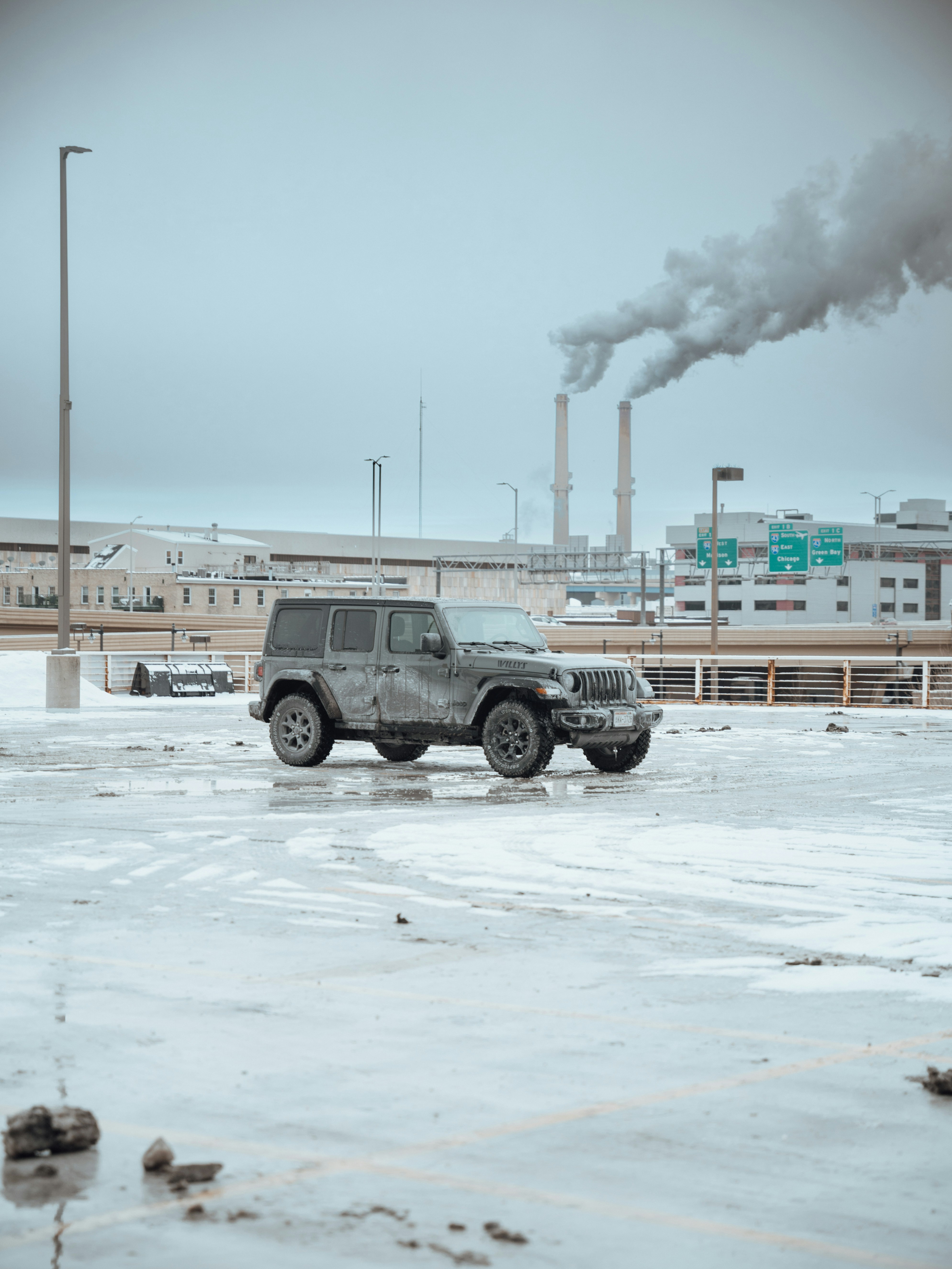 A rugged jeep parked on a snowy surface, with smoke billowing from distant chimneys, reflecting a blend of nature and urban industry.