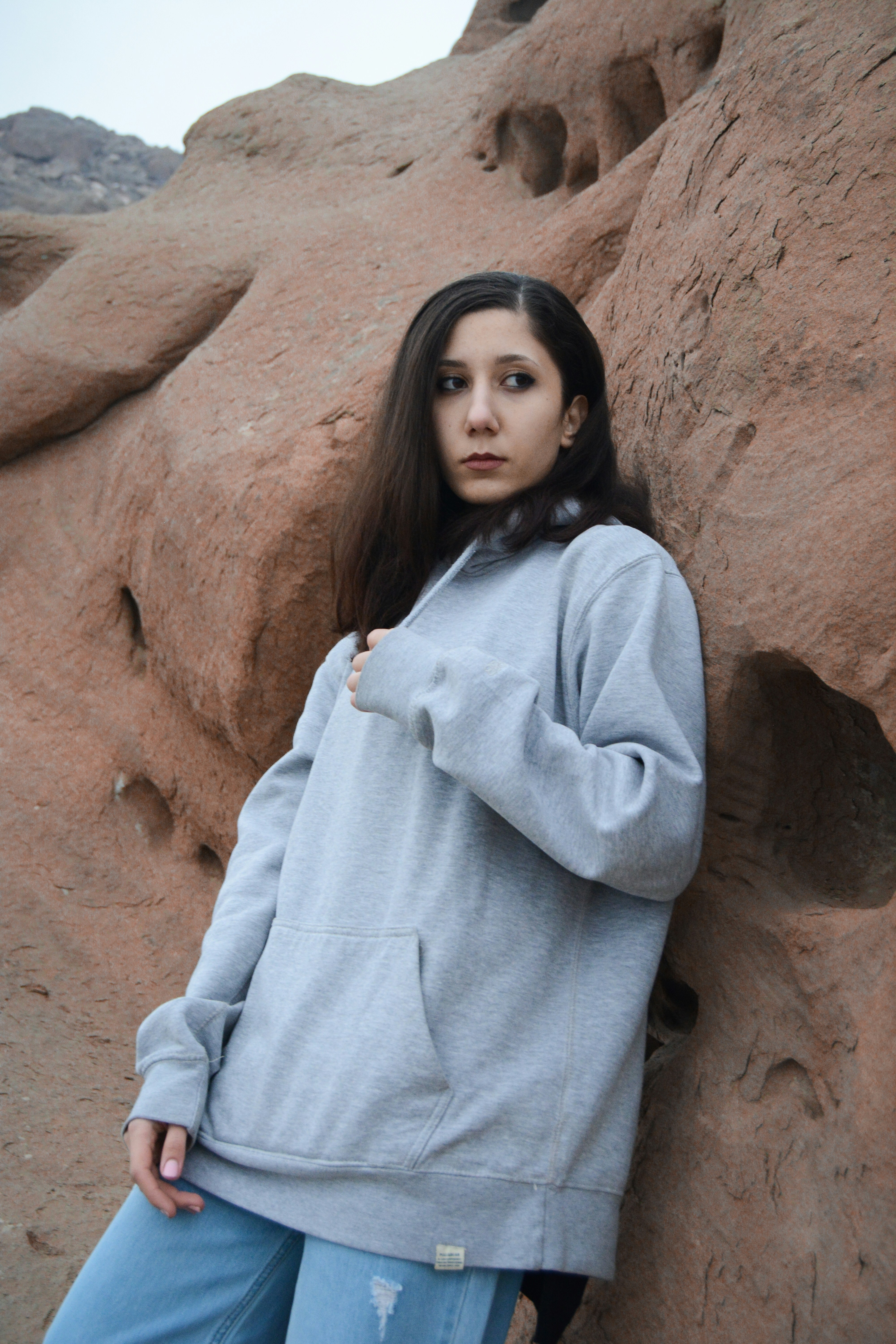 A young woman in a gray hoodie stands pensively against a rocky backdrop, blending casual attire with the ruggedness of the landscape.