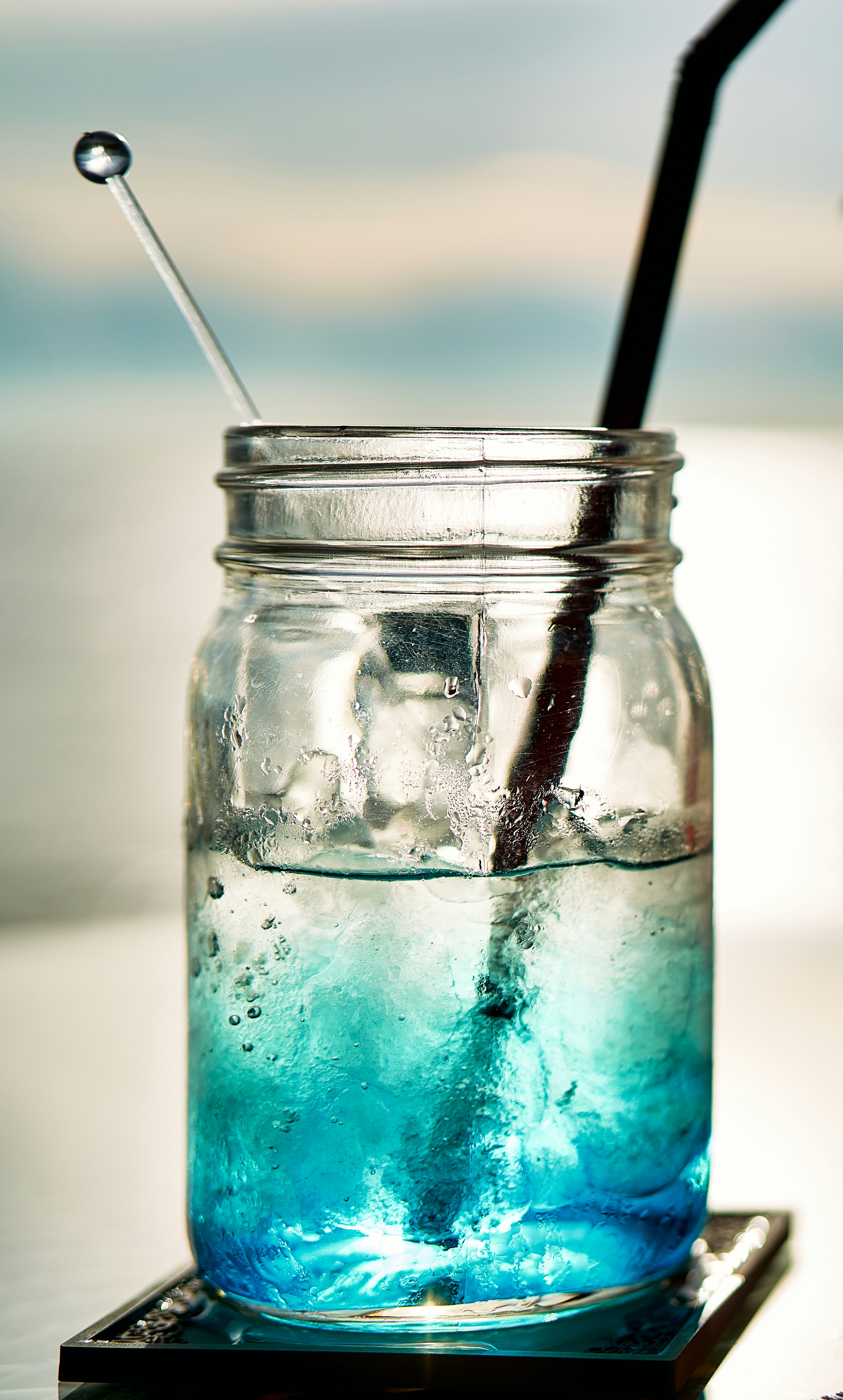 Vibrant blue beverage in a glass jar, with condensation and bubbles, set against a blurred background of water and sky.