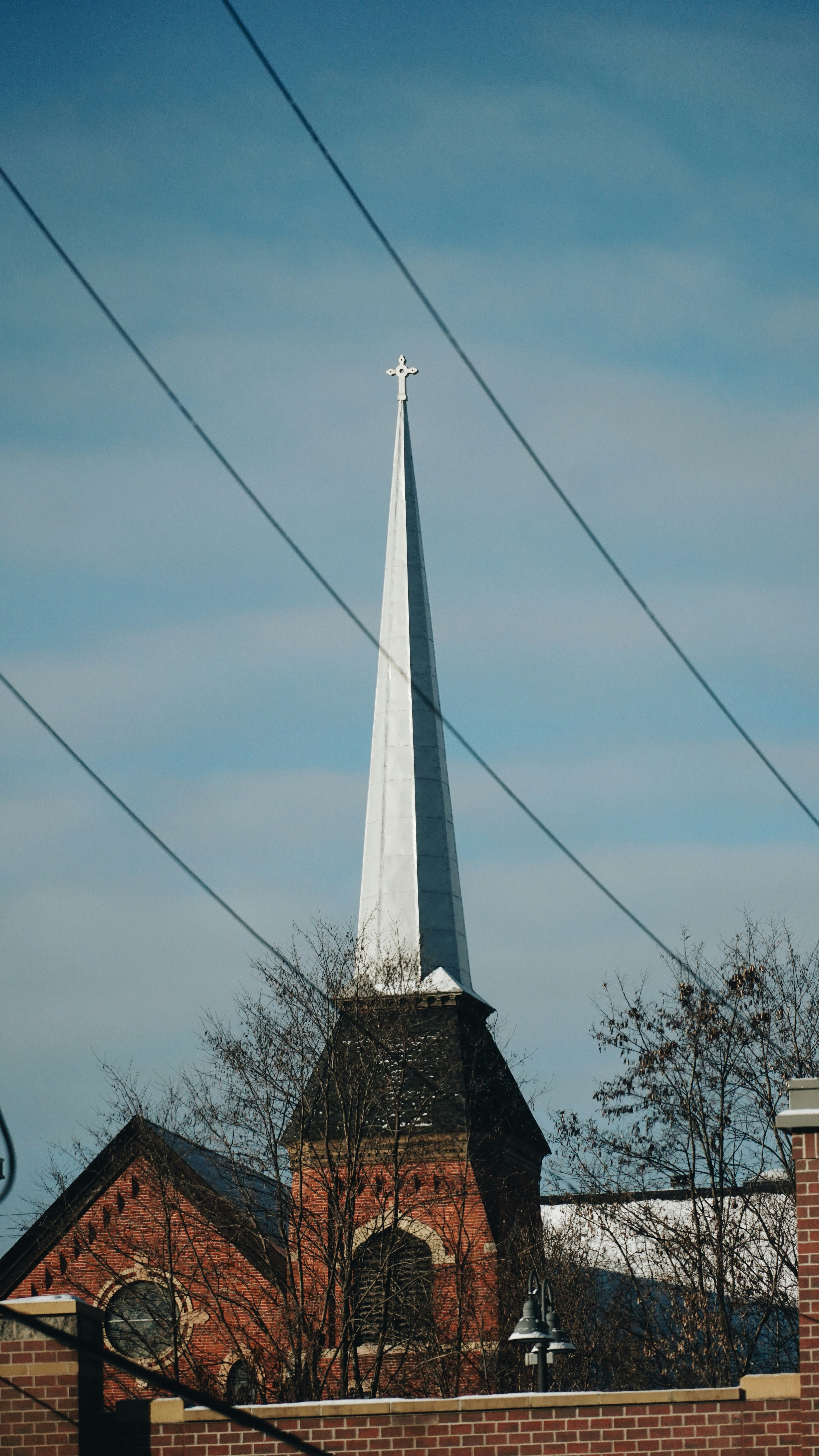 A tall church steeple rises against a clear blue sky, topped with a cross, framed by bare tree branches and power lines.