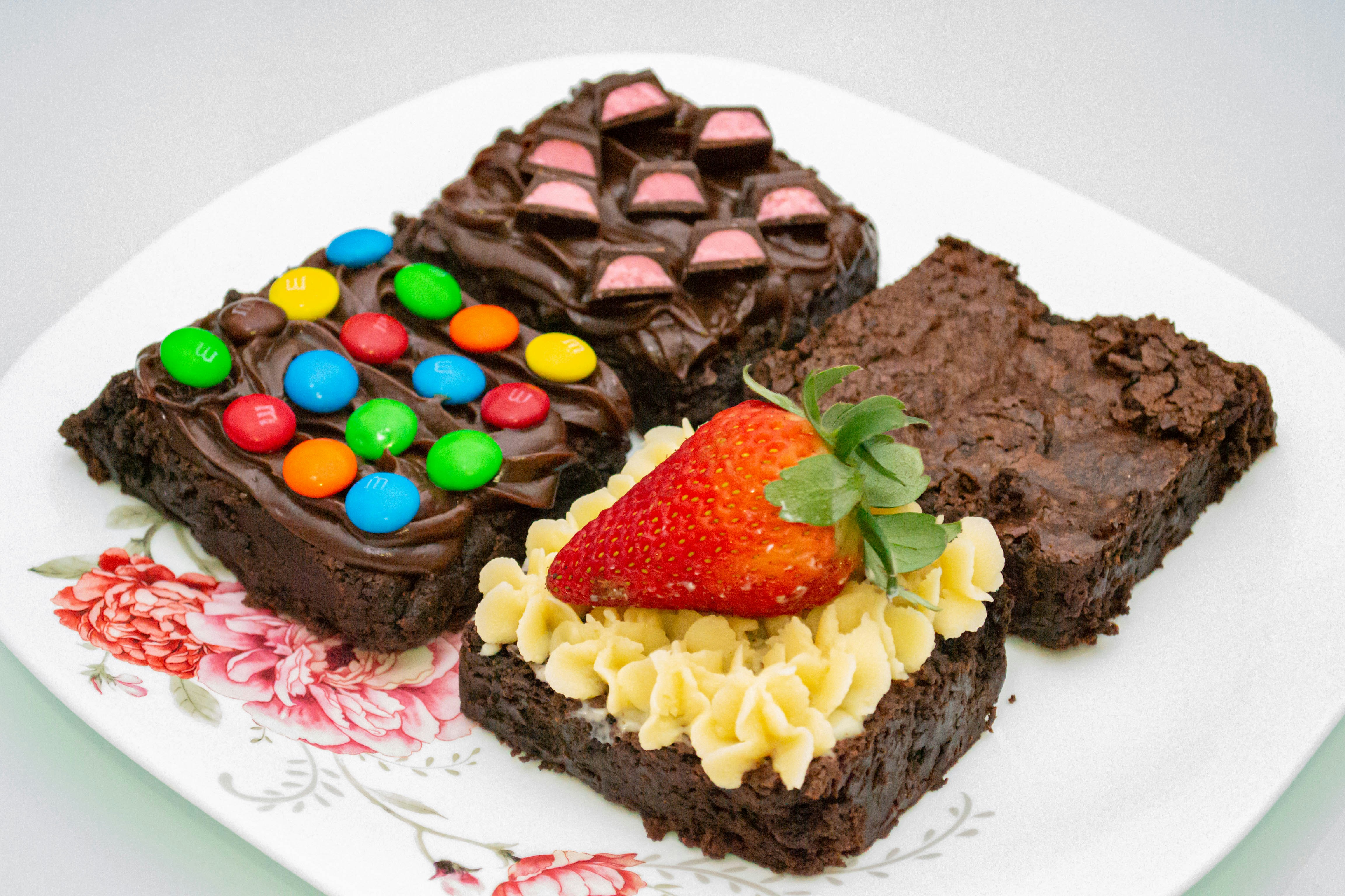 Four distinct brownies topped with colorful candies and a fresh strawberry, elegantly arranged on a floral plate.
