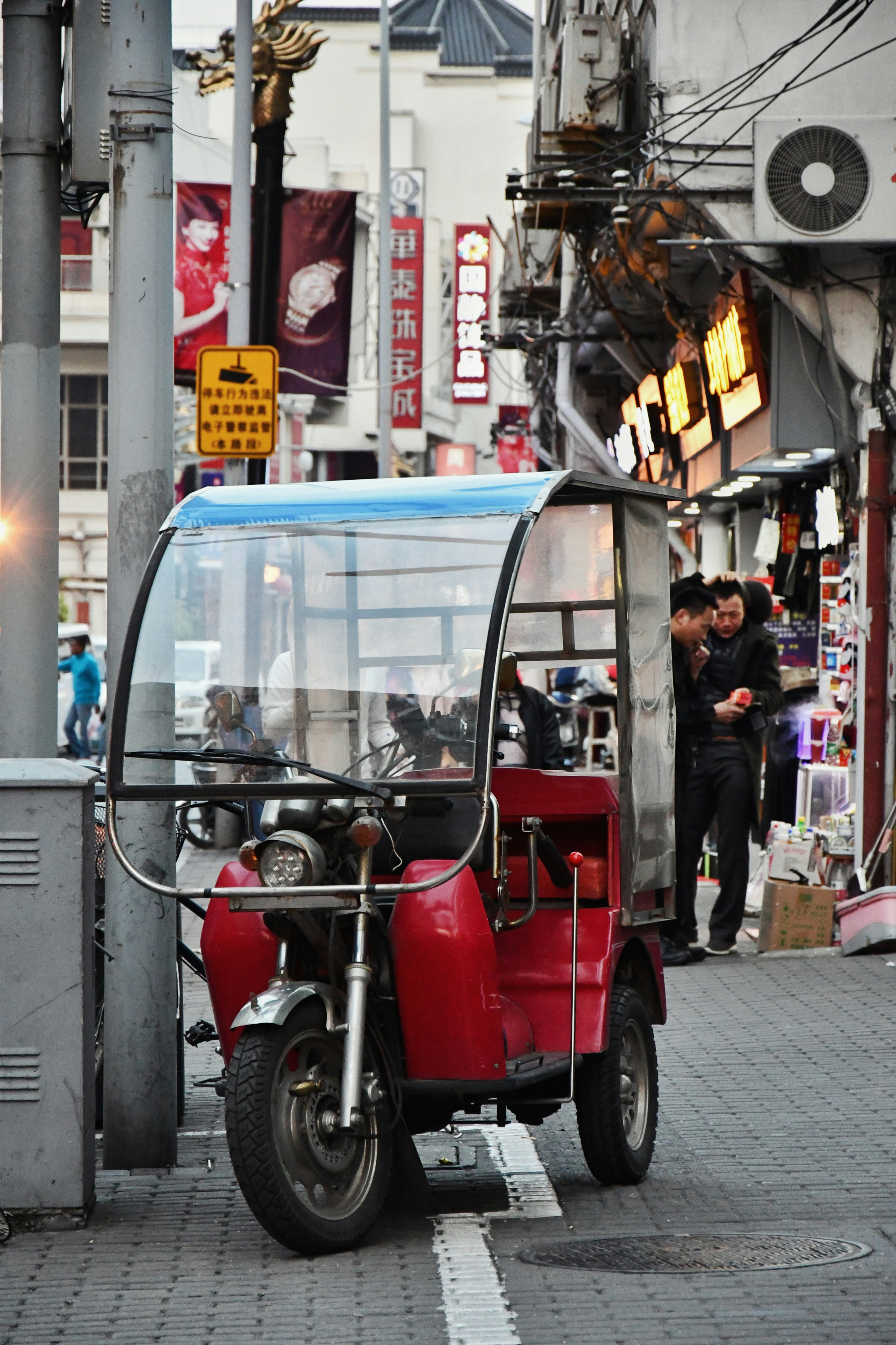 A vibrant red three-wheeled vehicle parked on a bustling street, surrounded by shops and pedestrians in an urban setting.
