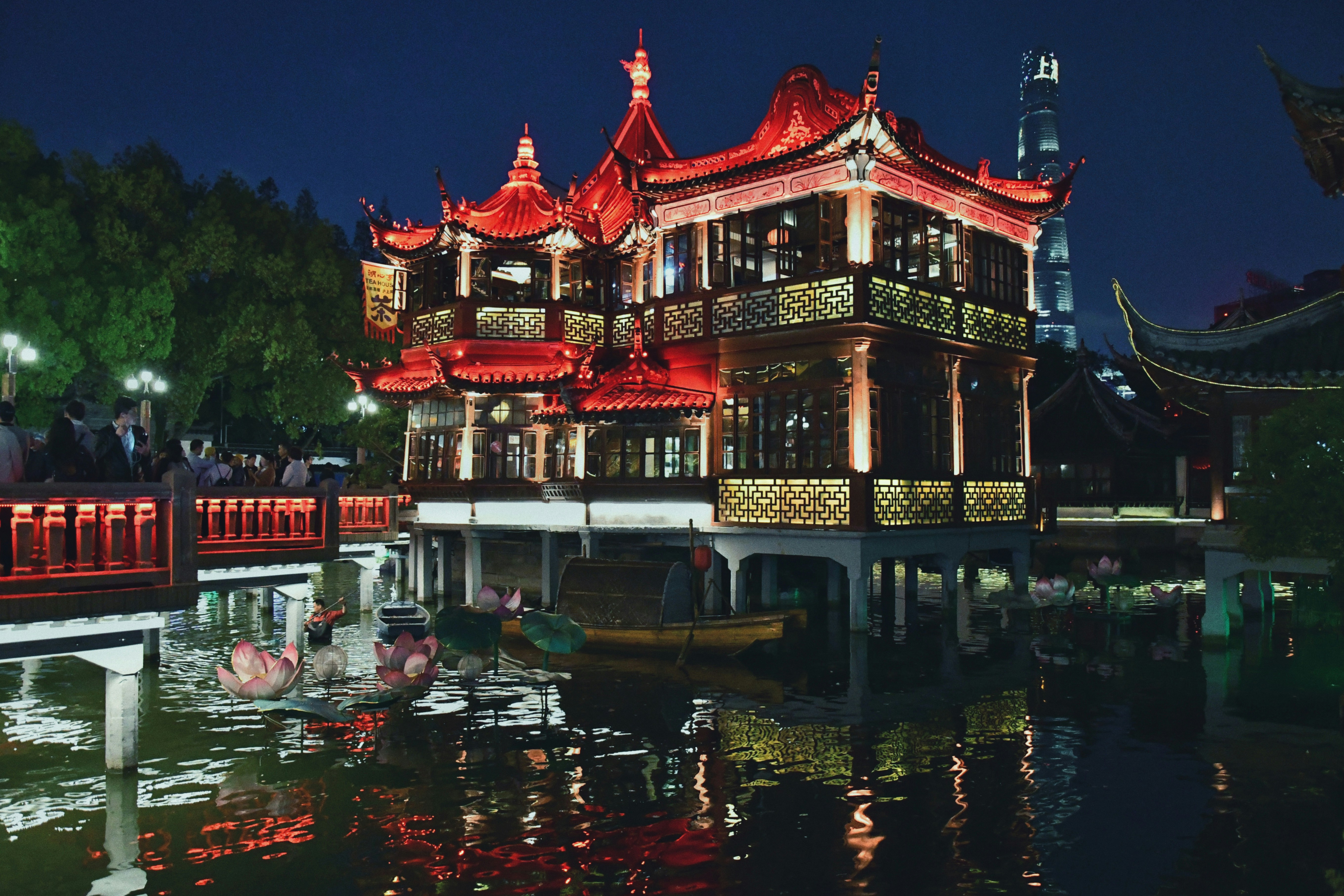 Traditional Chinese pavilion illuminated in red lights, reflecting in the calm waters at night, surrounded by lush greenery and visitors.