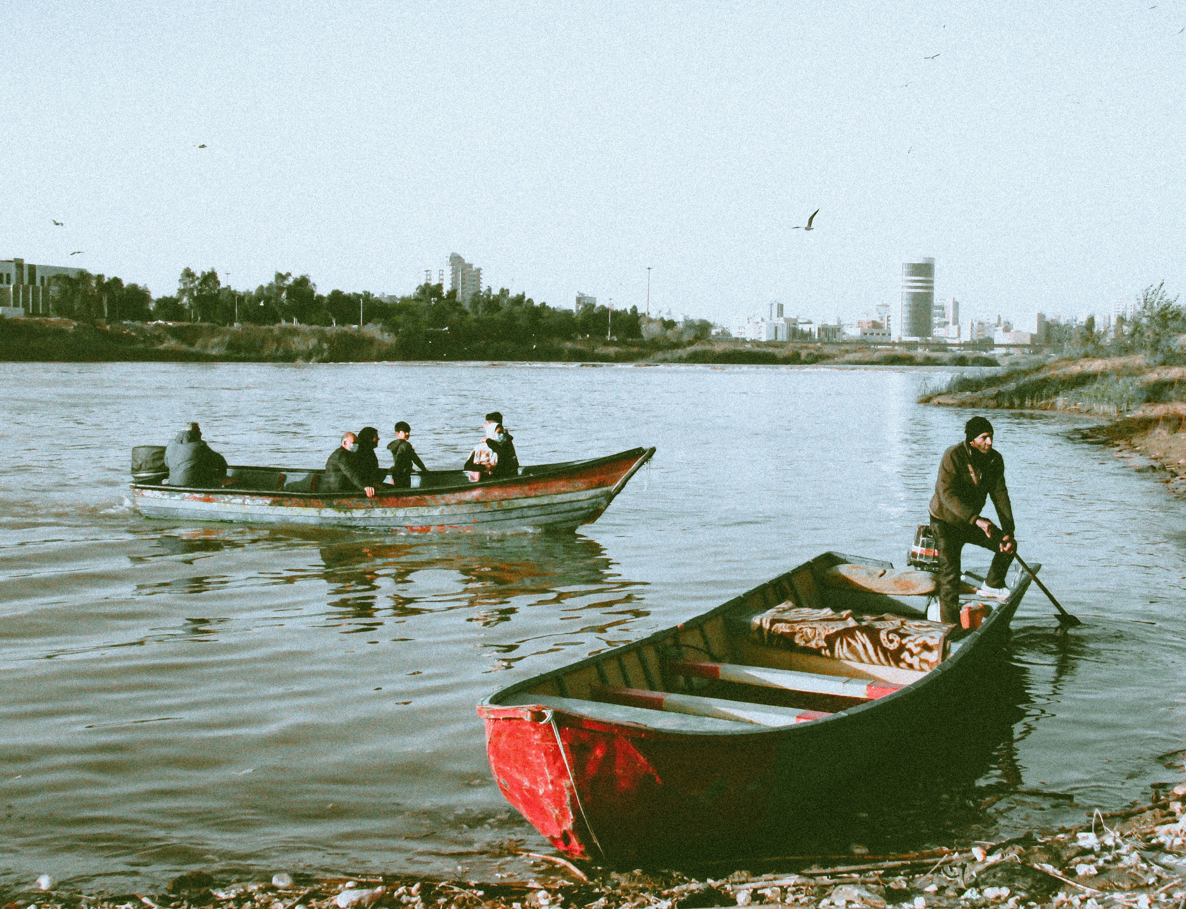 Two boats navigate a serene river, with individuals engaged in conversation and leisure activities against an urban backdrop. The tranquil scene captures the essence of community and relaxation.