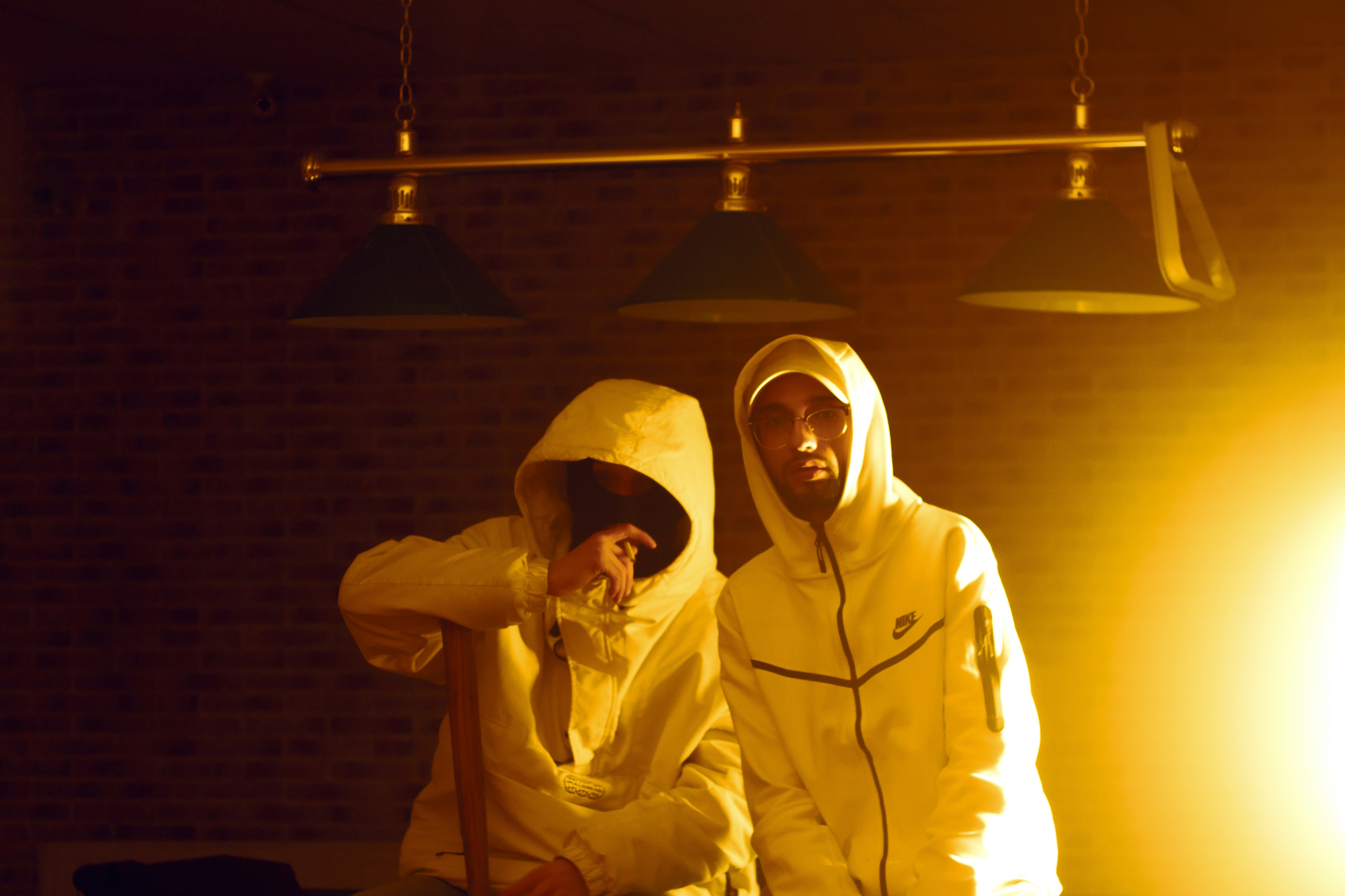 Hooded Duo in a Brick-Walled Studio Bathed in Ember Glow