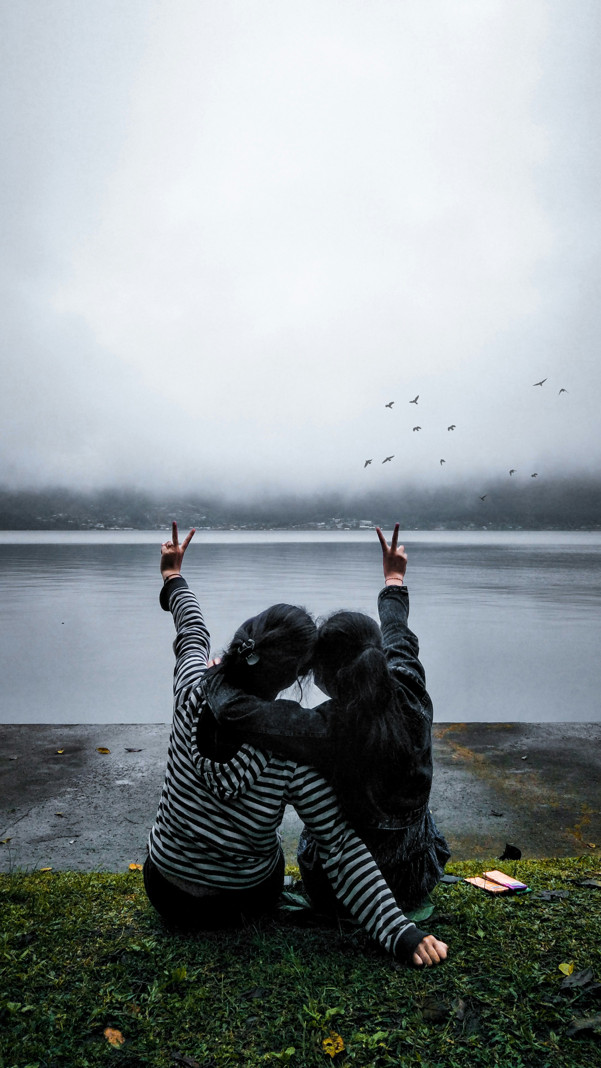 Two friends sitting side by side, making peace signs against a misty lake backdrop. The serene atmosphere captures a moment of friendship and tranquility.