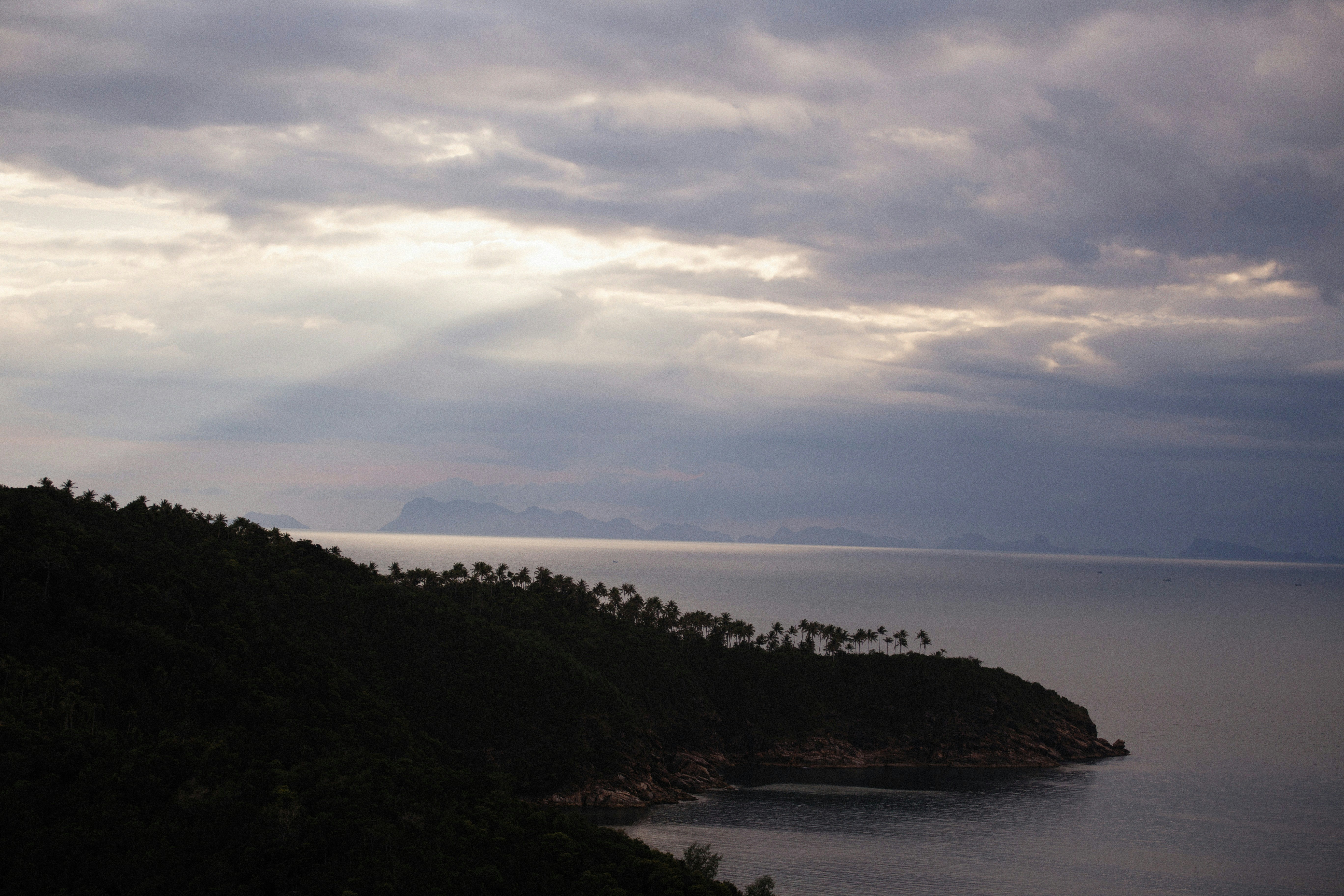 A tranquil coastal landscape featuring a lush green hillside meeting the calm sea under a moody sky. Rays of light break through the clouds, highlighting the serene atmosphere.