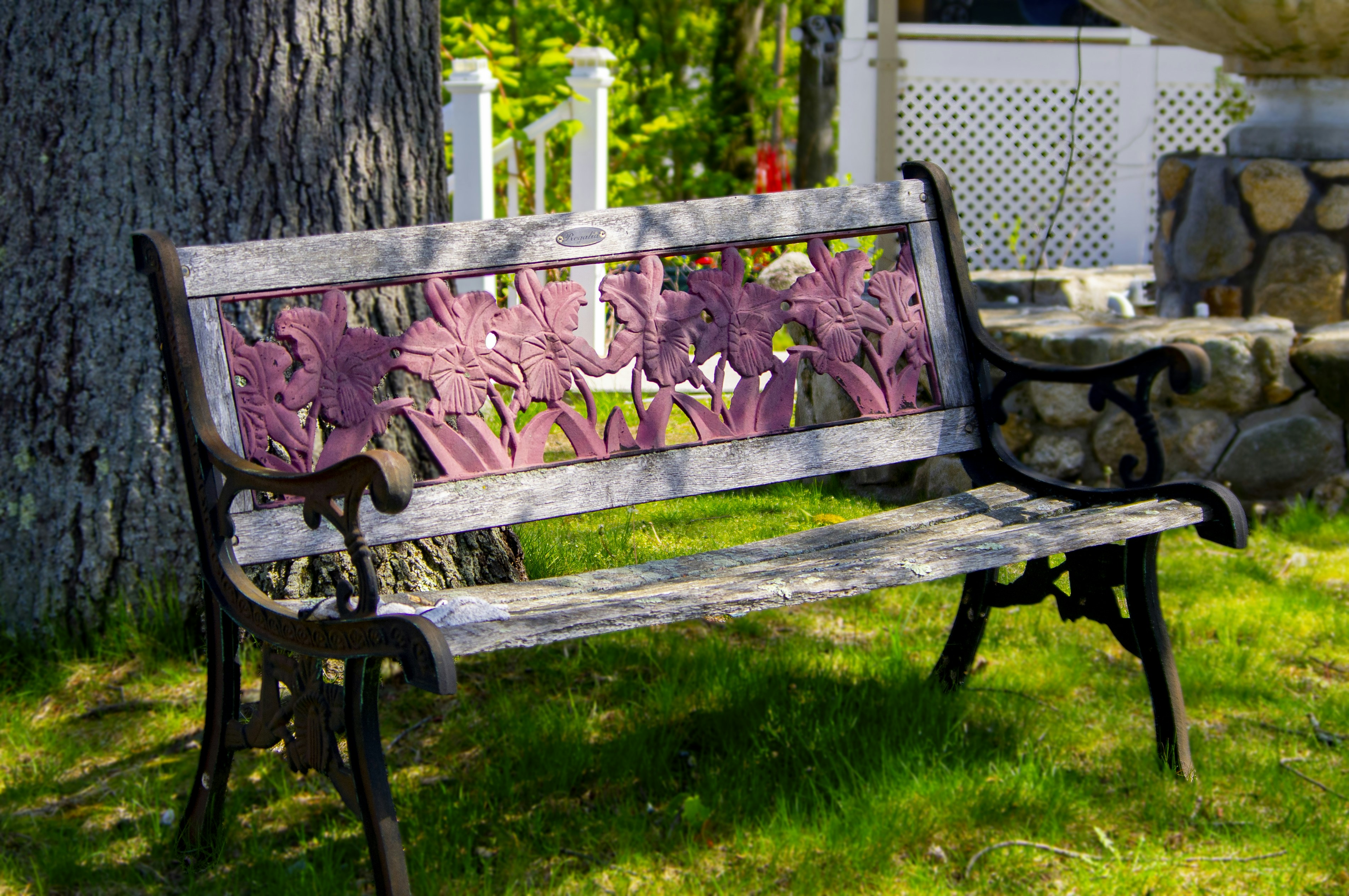 Ornate bench featuring floral designs, nestled in a lush garden setting under a tree. The scene invites relaxation and reflection.