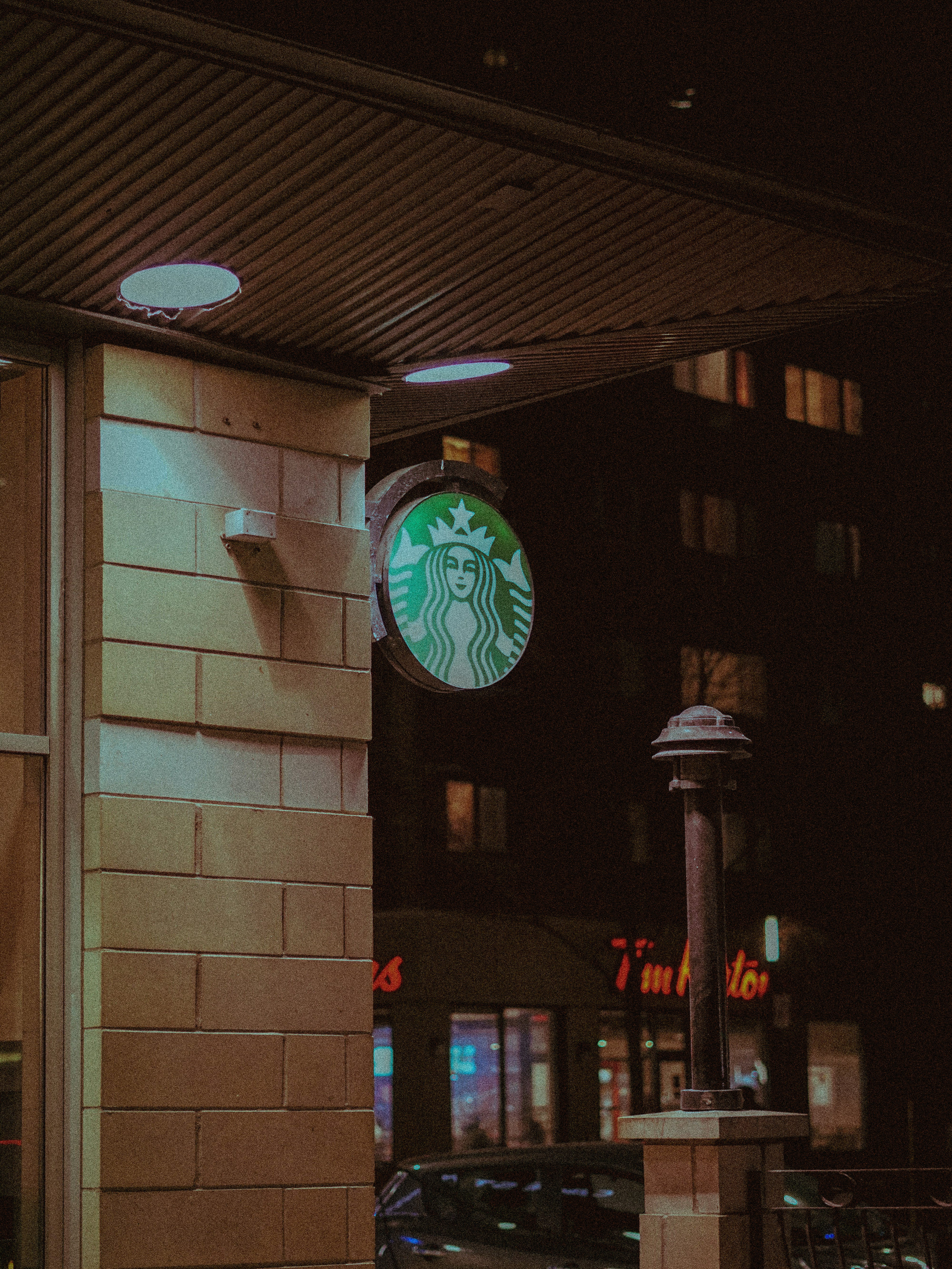 Illuminated Starbucks logo on a building corner, with warm lighting contrasting against the night. Tim Hortons sign visible in the background.