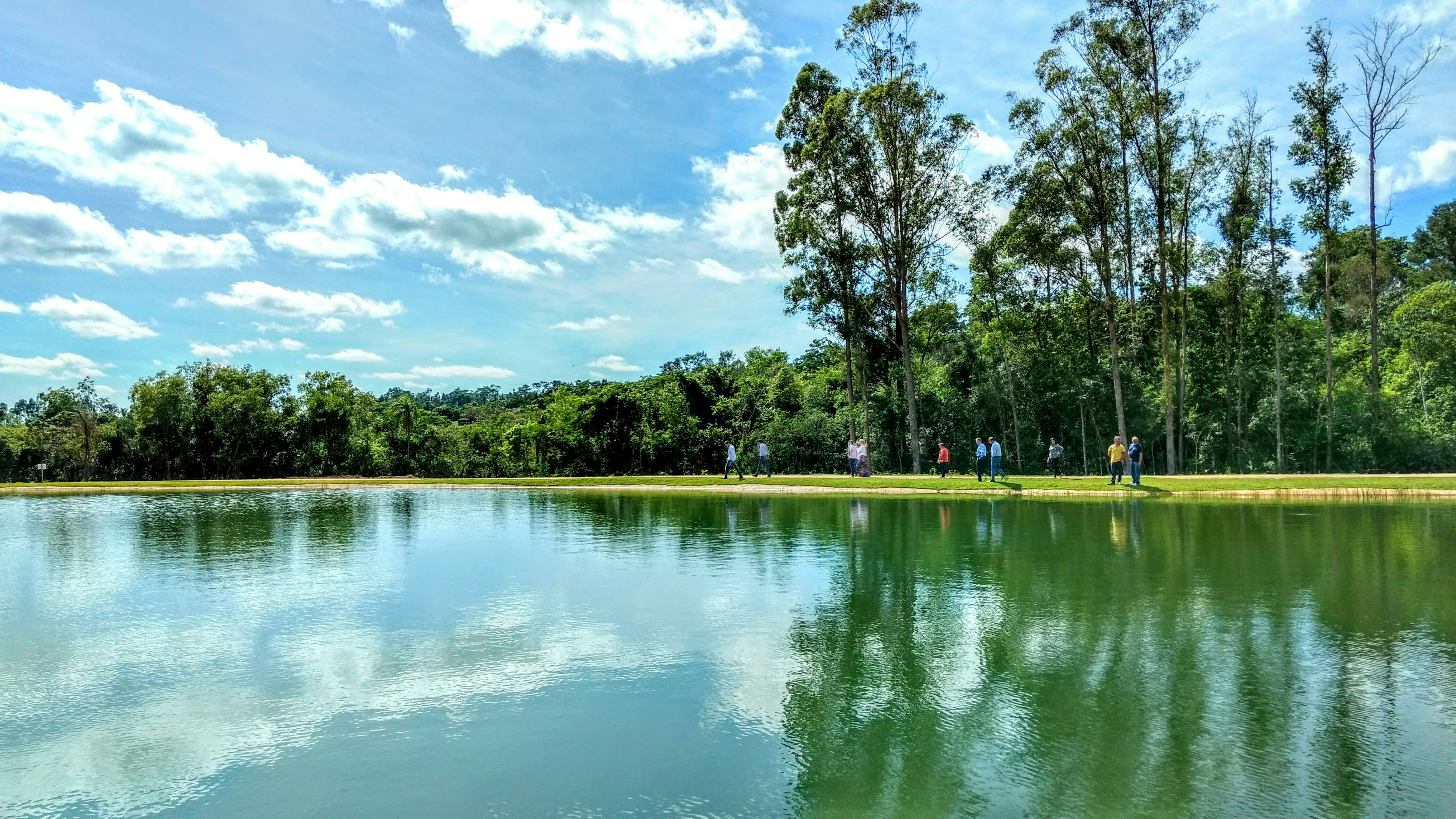 A photograph of a calm lake with a tree-lined shore and a distant row of walkers along the far bank. The reflections on the water emphasize the tranquil landscape.