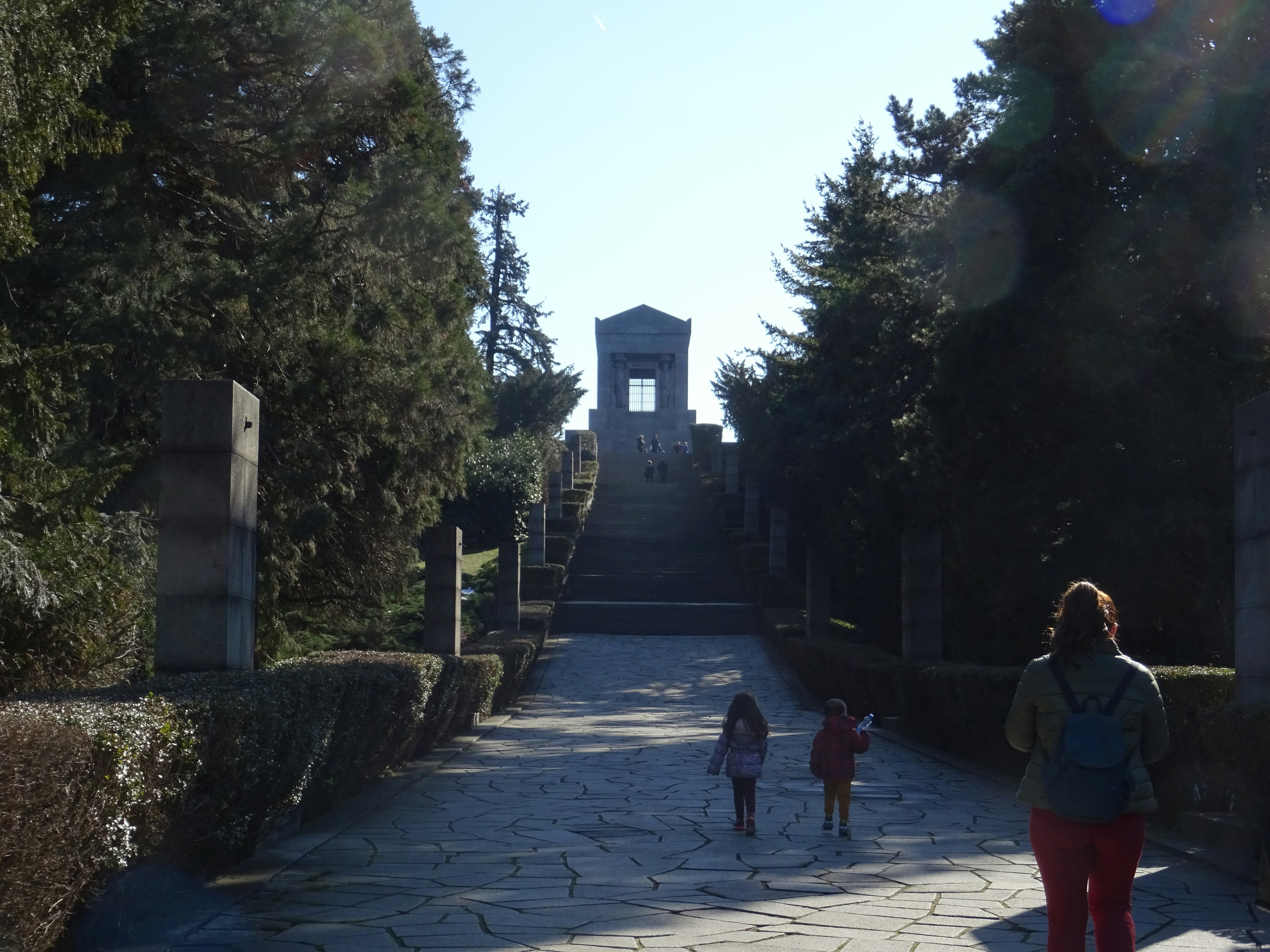 Shaded pathway leading to a distant monument under a clear sky, flanked by tall trees and stone pillars.