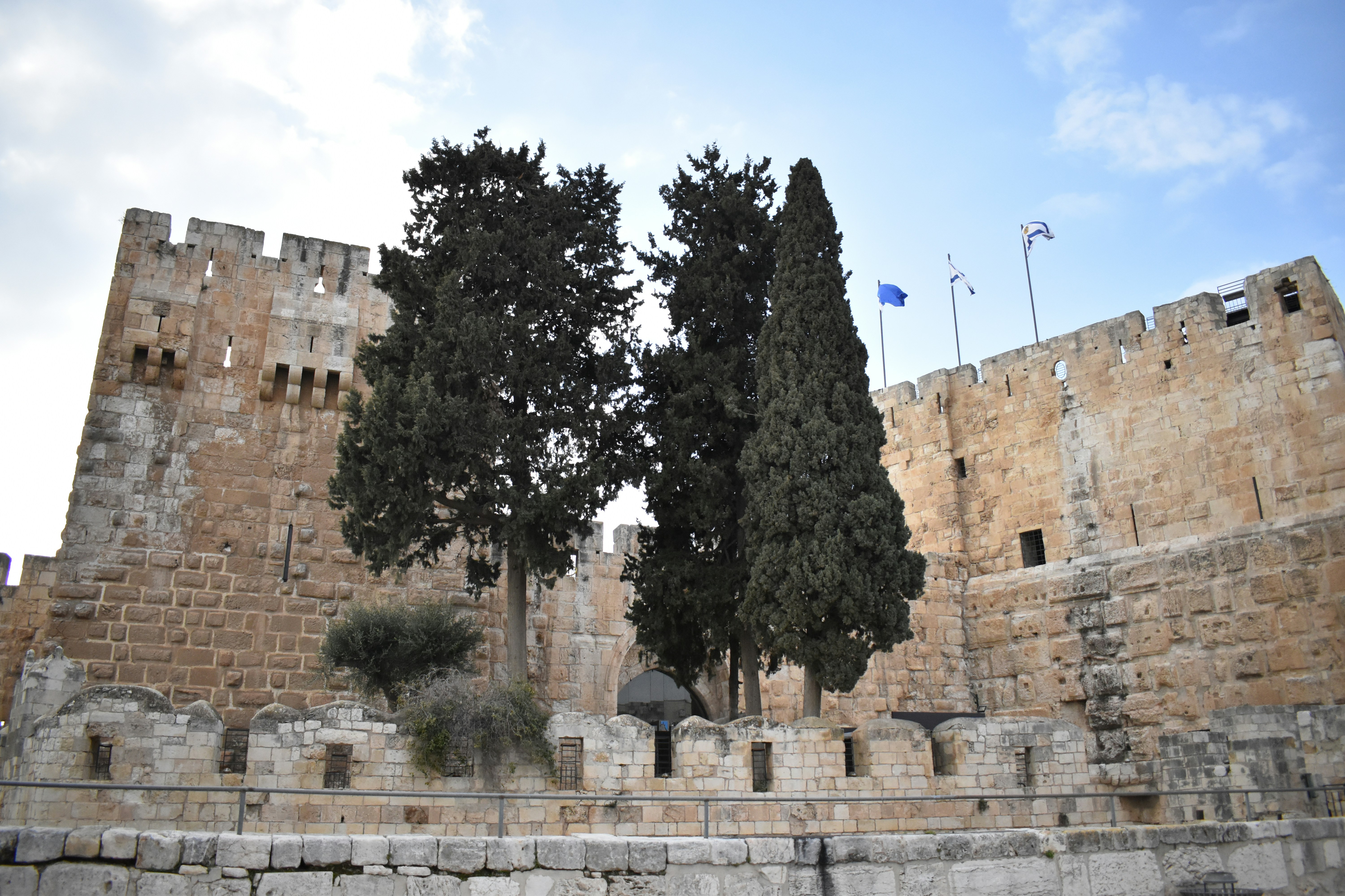 Stone castle with two tall trees in front, set against a partly cloudy sky.
