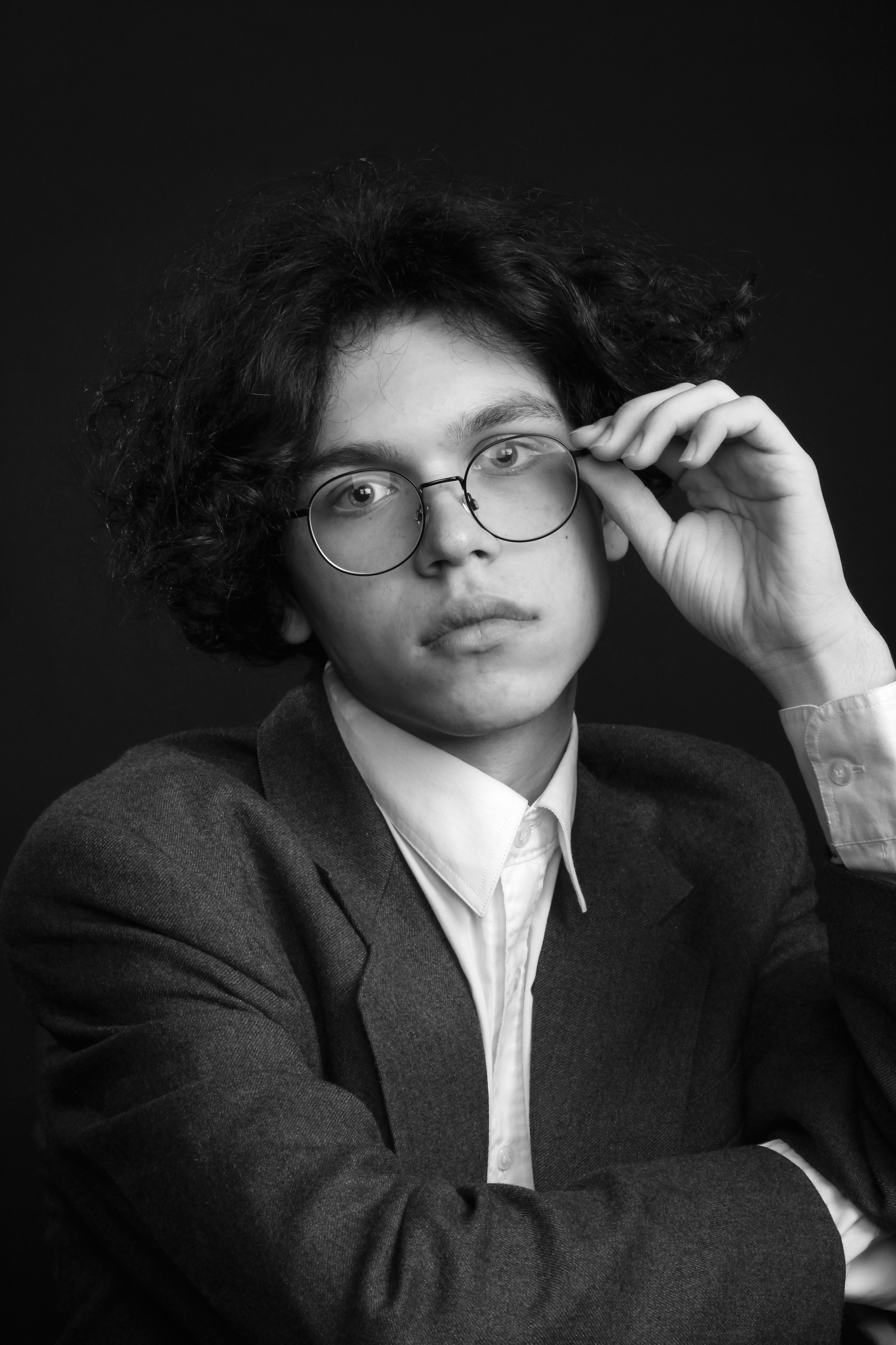 Young man in a tailored suit with round glasses, striking a contemplative pose against a dark background.