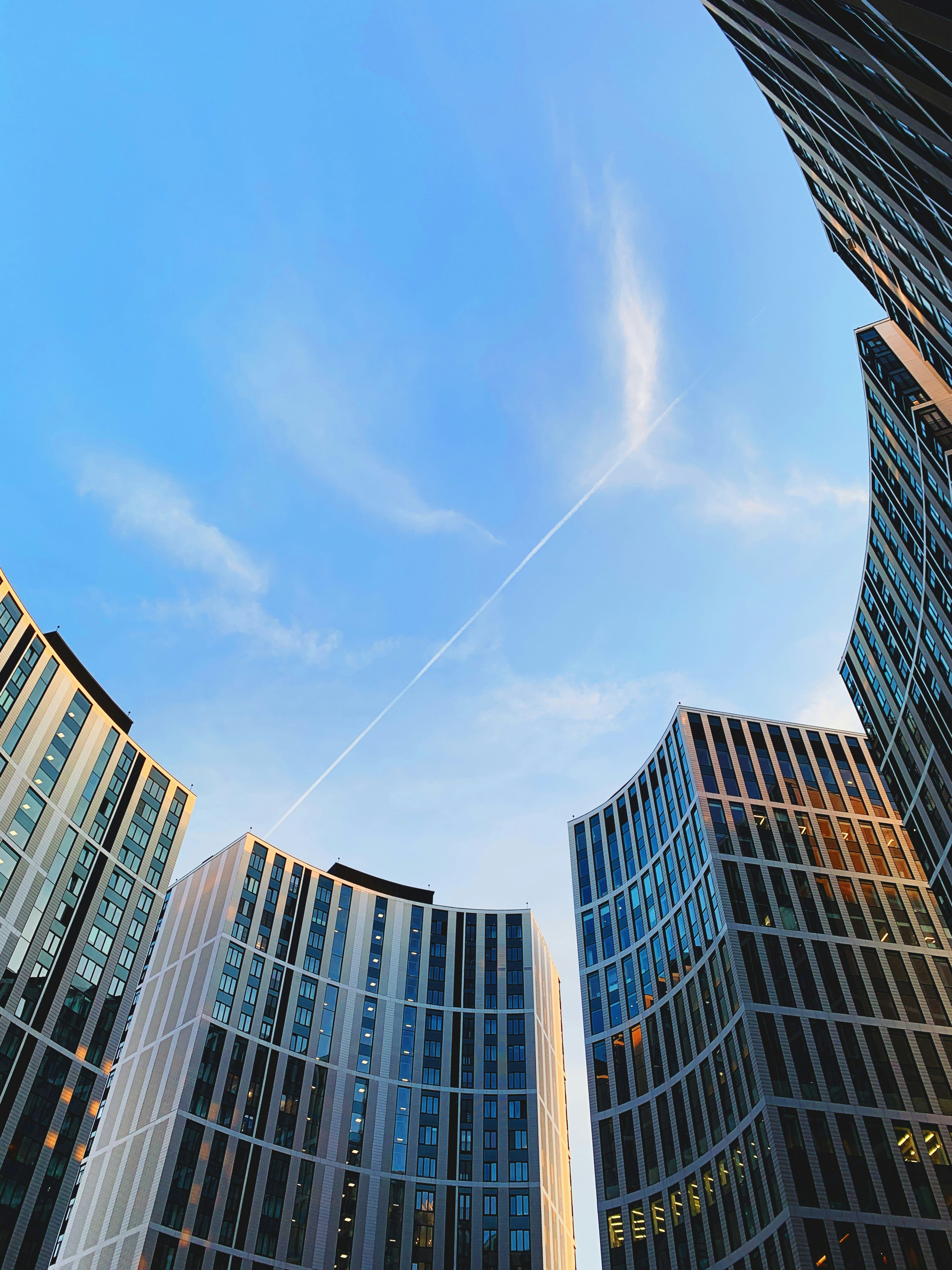 Convergence Under a Skyward Arc: Glass Towers Framed by a Diagonal Contrail