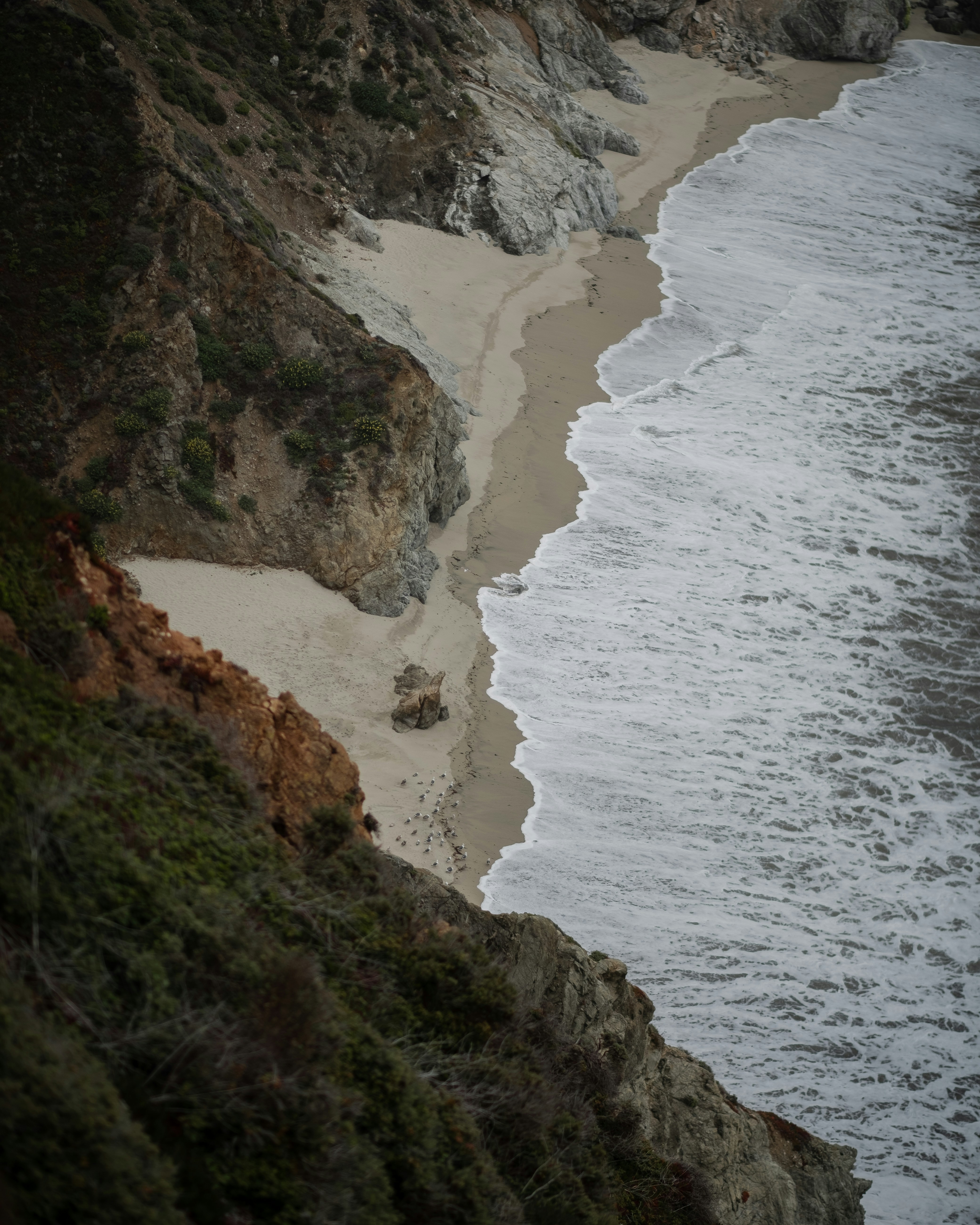 Waves gently lap at a secluded beach framed by rugged cliffs, with hints of vegetation adding texture to the scene.