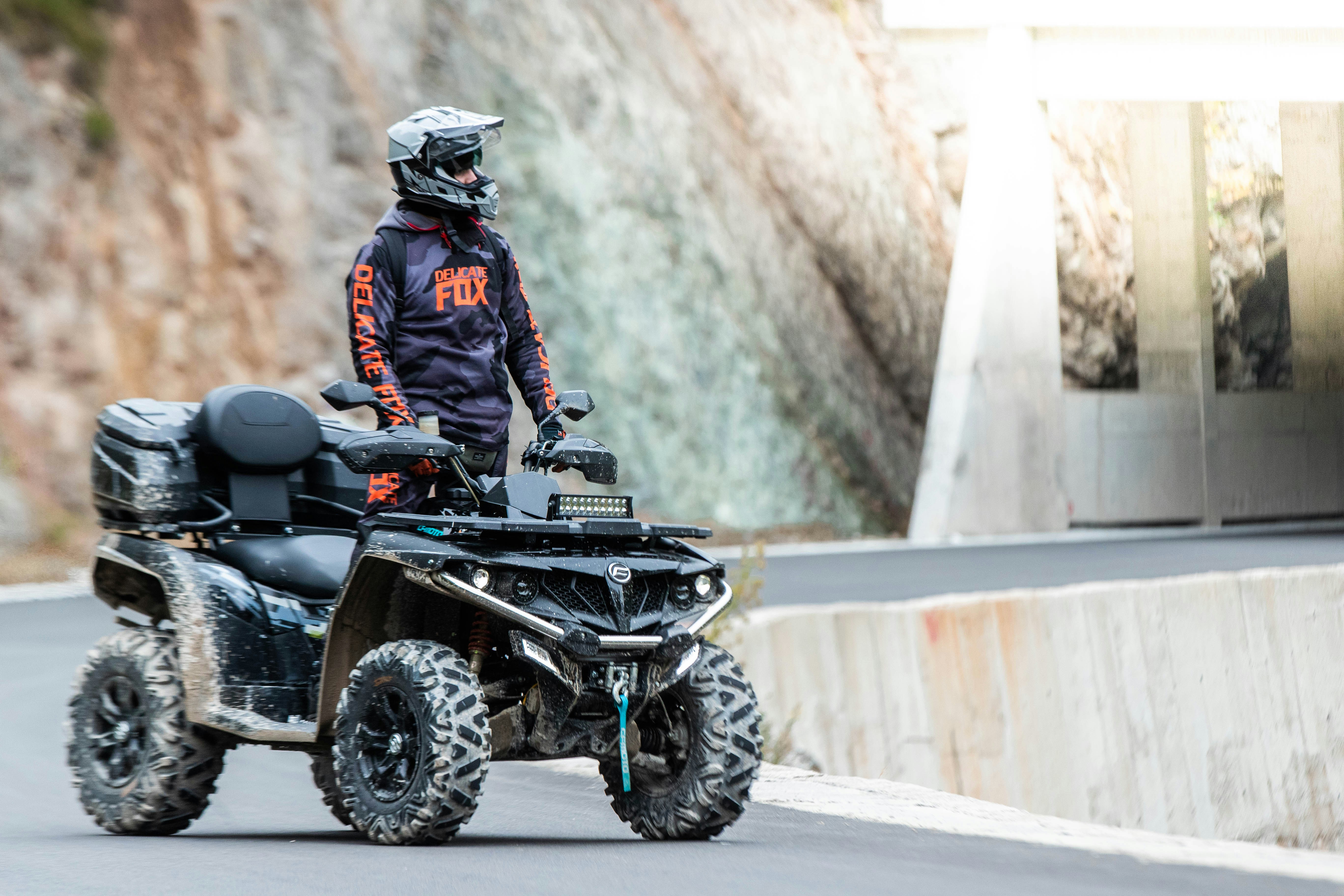 Person on an ATV with rugged gear, paused on a mountain road near a concrete tunnel.