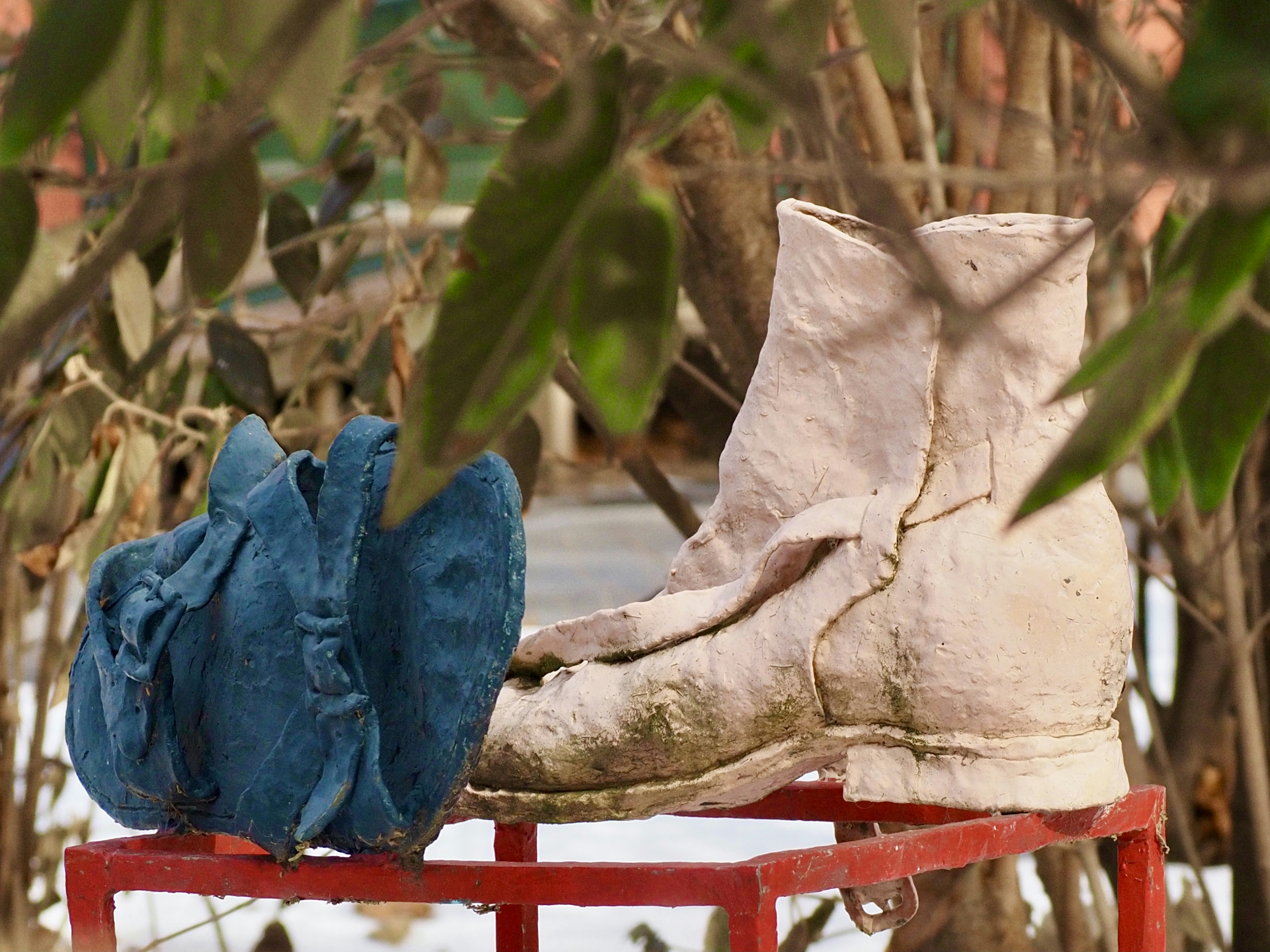 Two artistic shoe sculptures, one blue and one white, sit atop a red stand surrounded by foliage. Their unique textures and colors create a striking contrast.