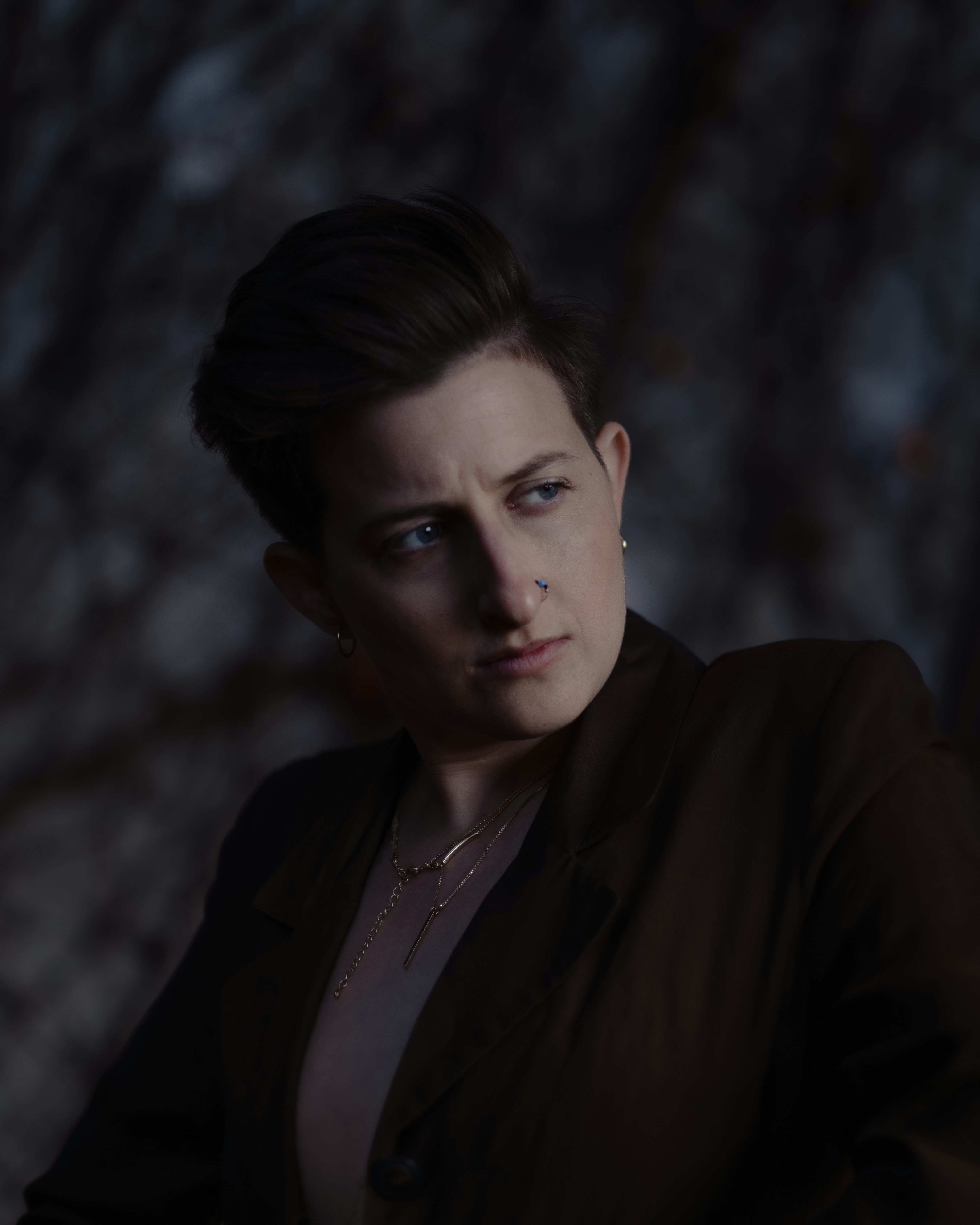 Individual with short hair and layered jewelry gazes thoughtfully against a textured backdrop.