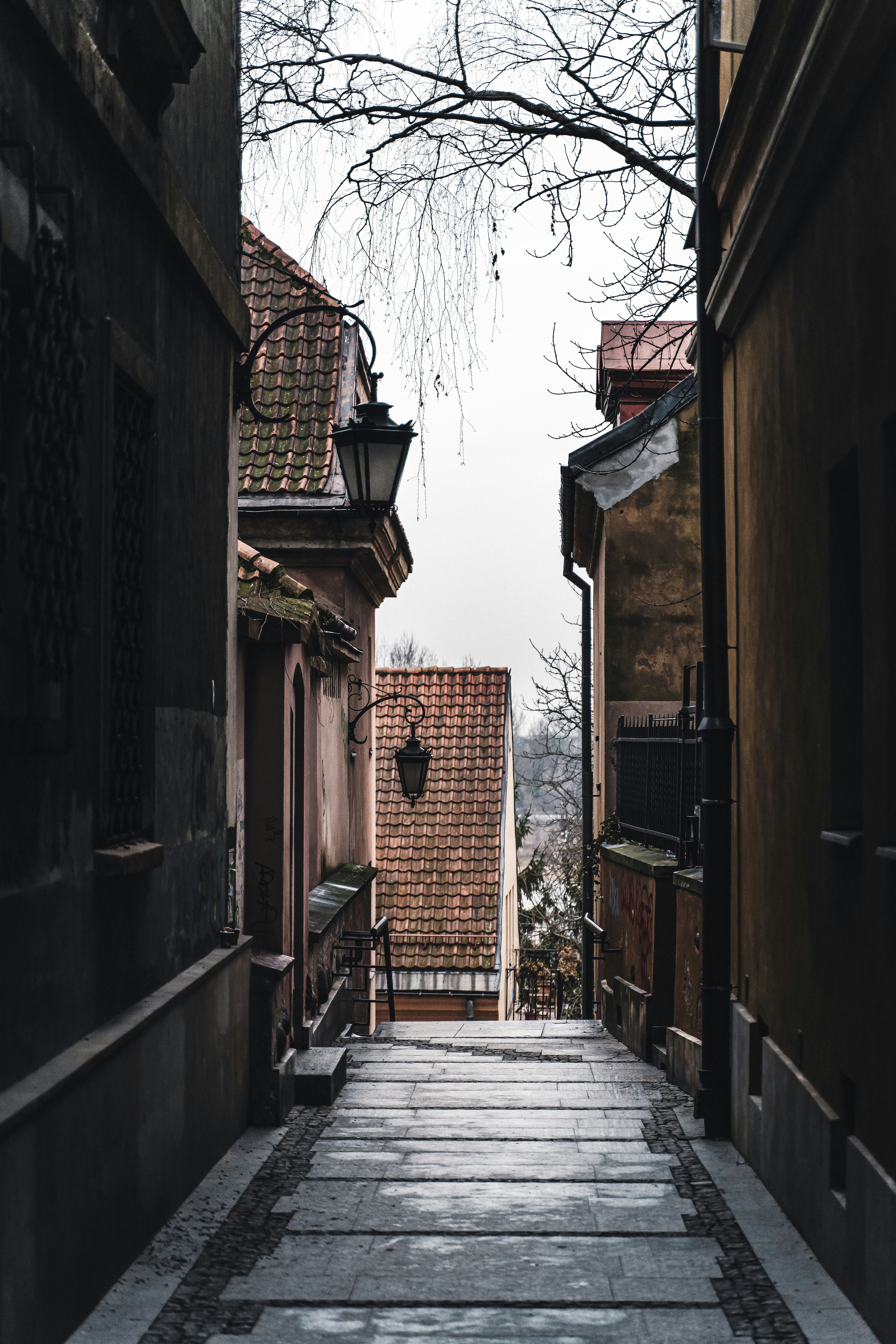 Narrow alleyway flanked by historic buildings, leading down to a distant view. A vintage lamp hangs above, illuminating the serene descent.