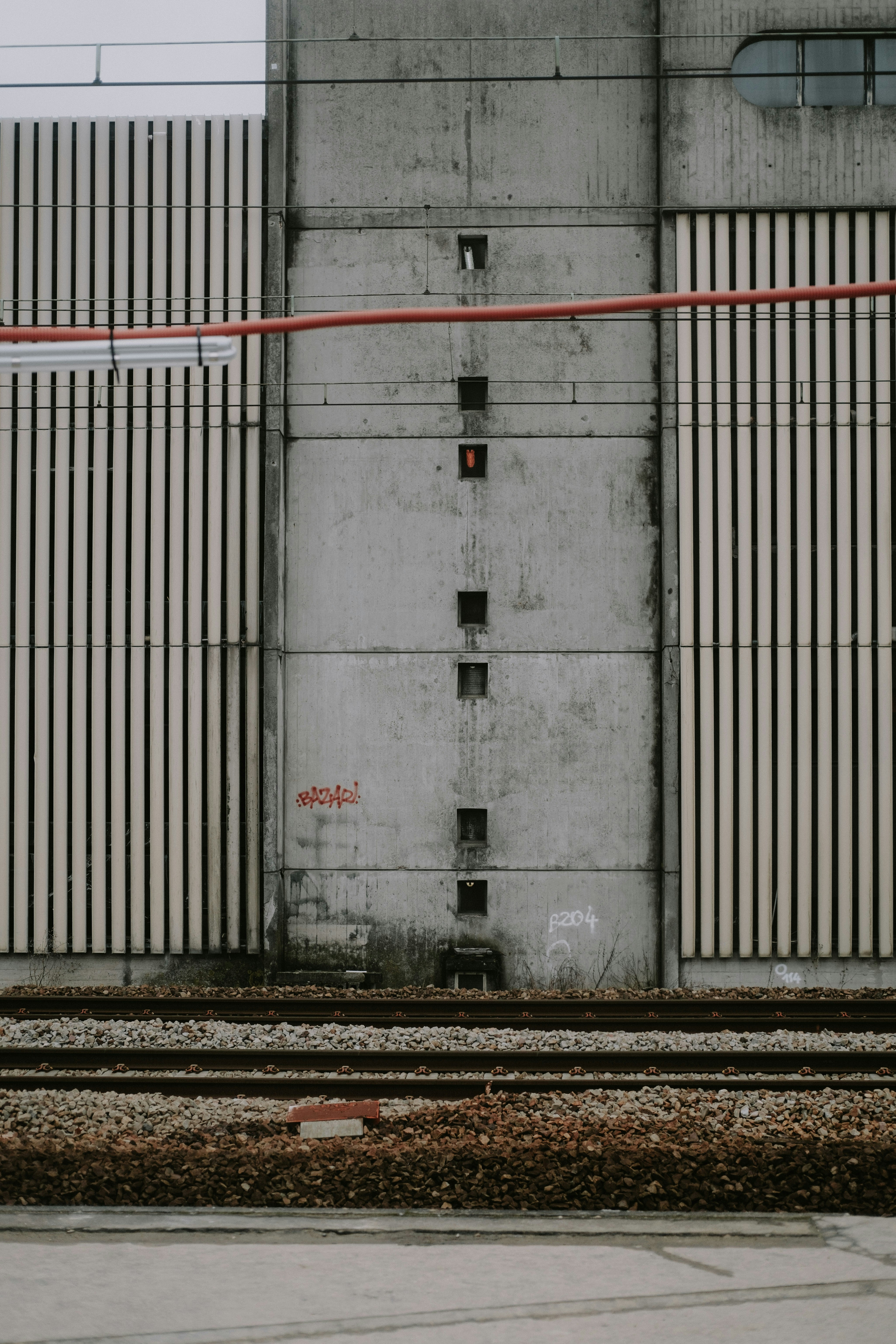 Textured concrete wall with vertical slats and subtle graffiti, set against railway tracks below.