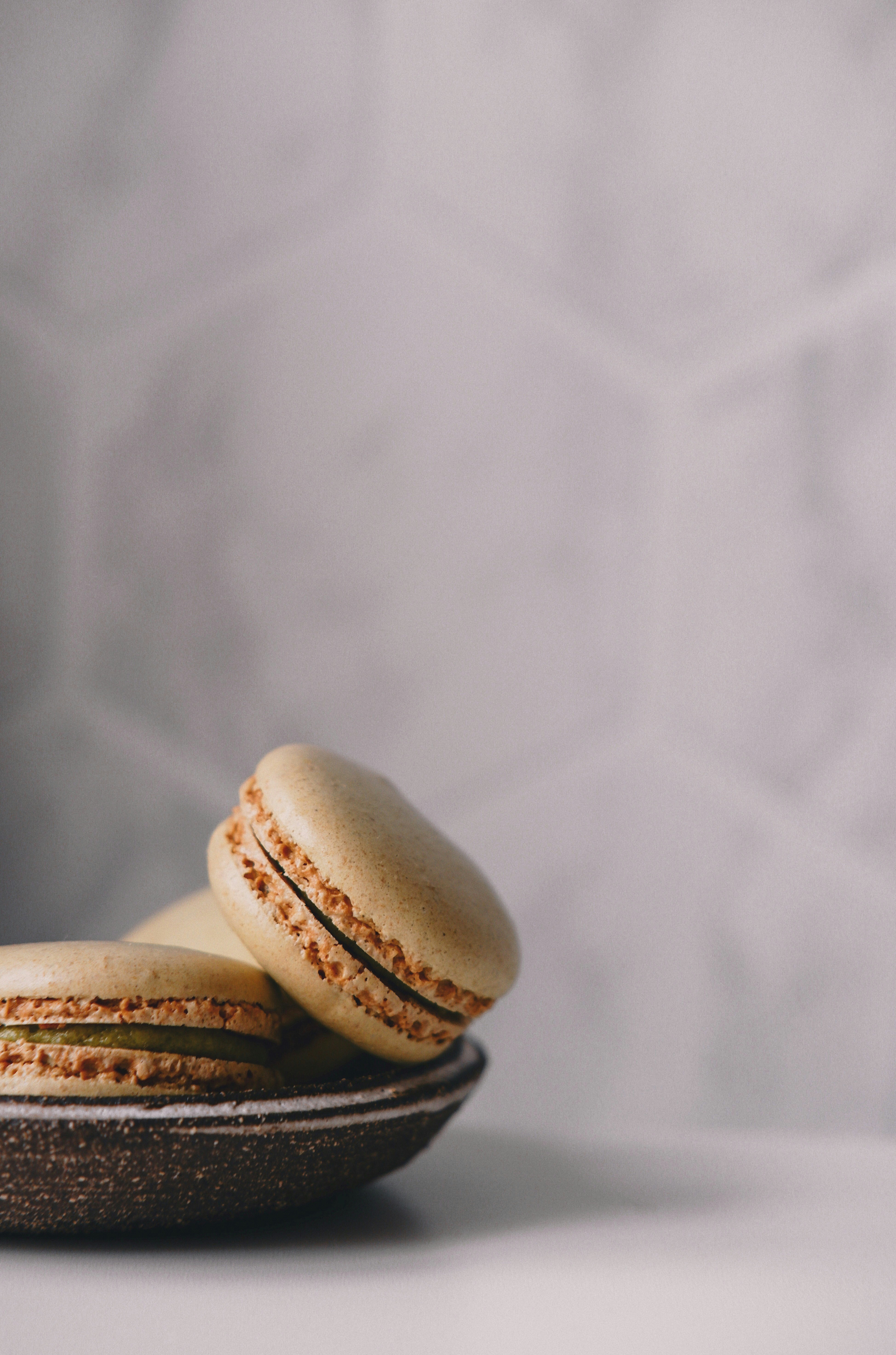 Two almond macarons rest on a dark ceramic plate against a soft, neutral backdrop. The minimalist setup highlights delicate texture and subtle color.
