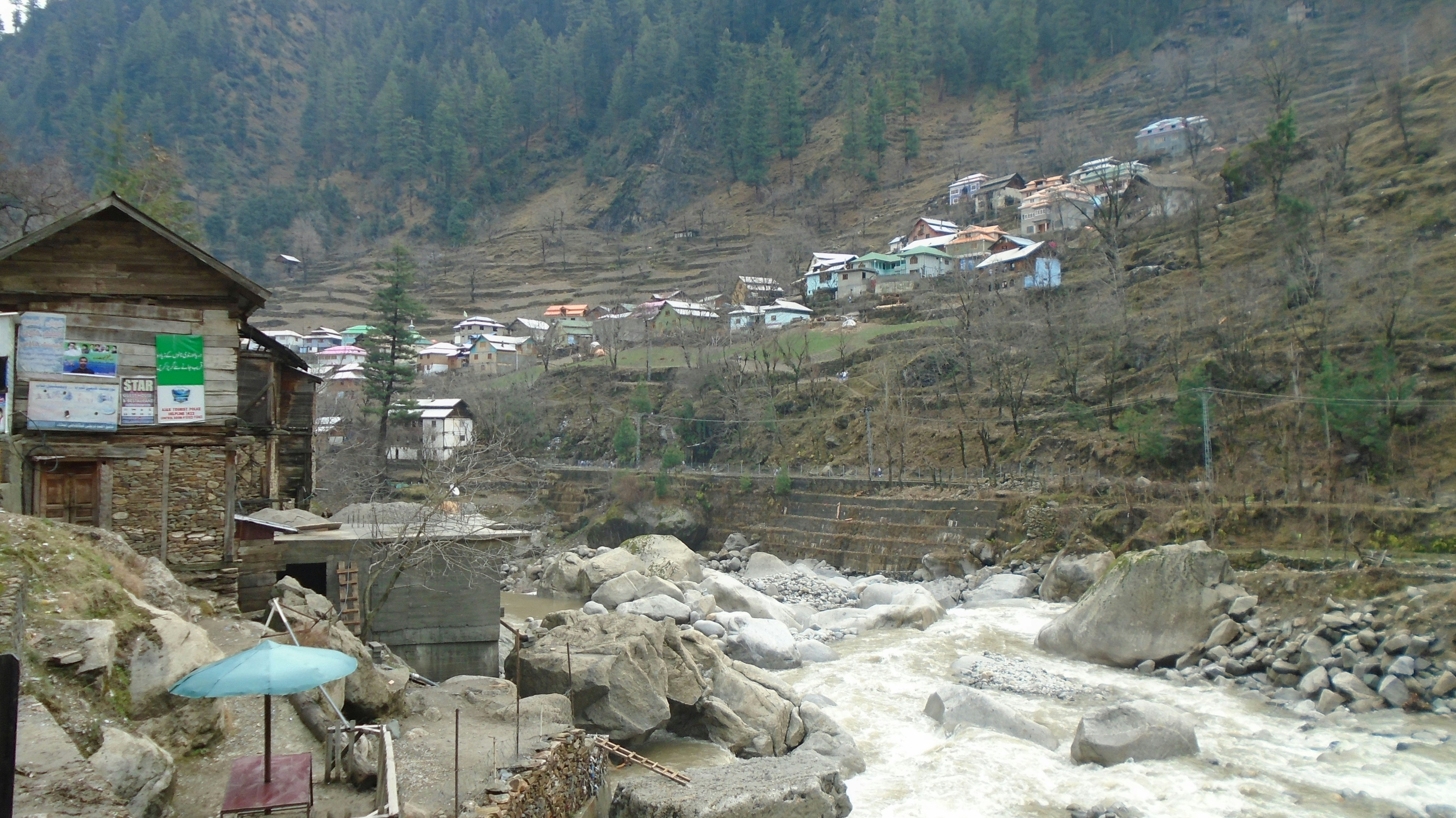 Rustic wooden structures line a rushing river, with colorful houses perched on a hillside in the background. The scene captures the essence of serene mountain living.