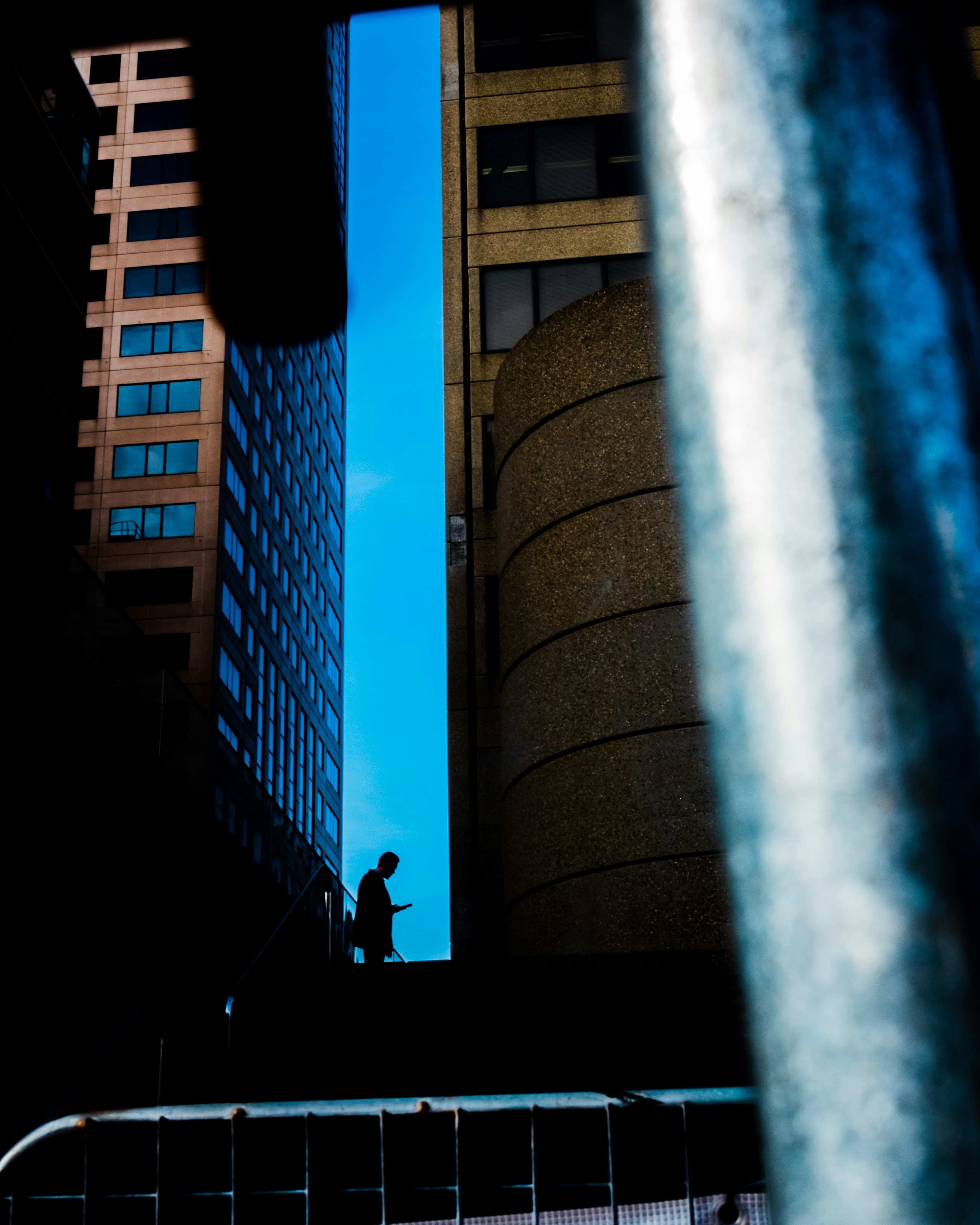 Silhouette of a person walking down stairs between towering buildings, framed by a bright blue sky. The urban landscape contrasts sharply with the subject's movement.