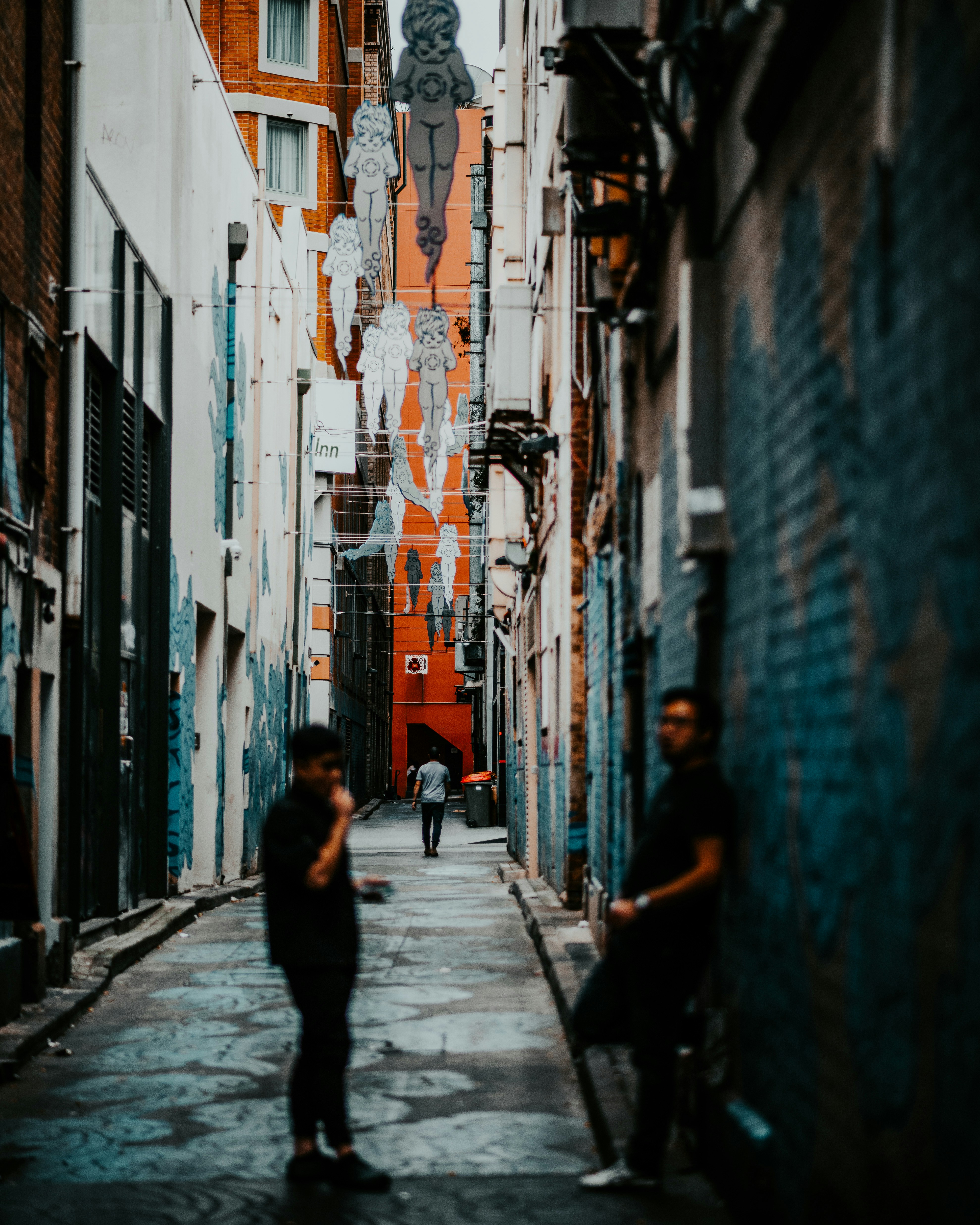 Vibrant street art decorates the walls of a narrow alley, where figures linger amidst the urban backdrop. A solitary figure walks towards the distant entrance.