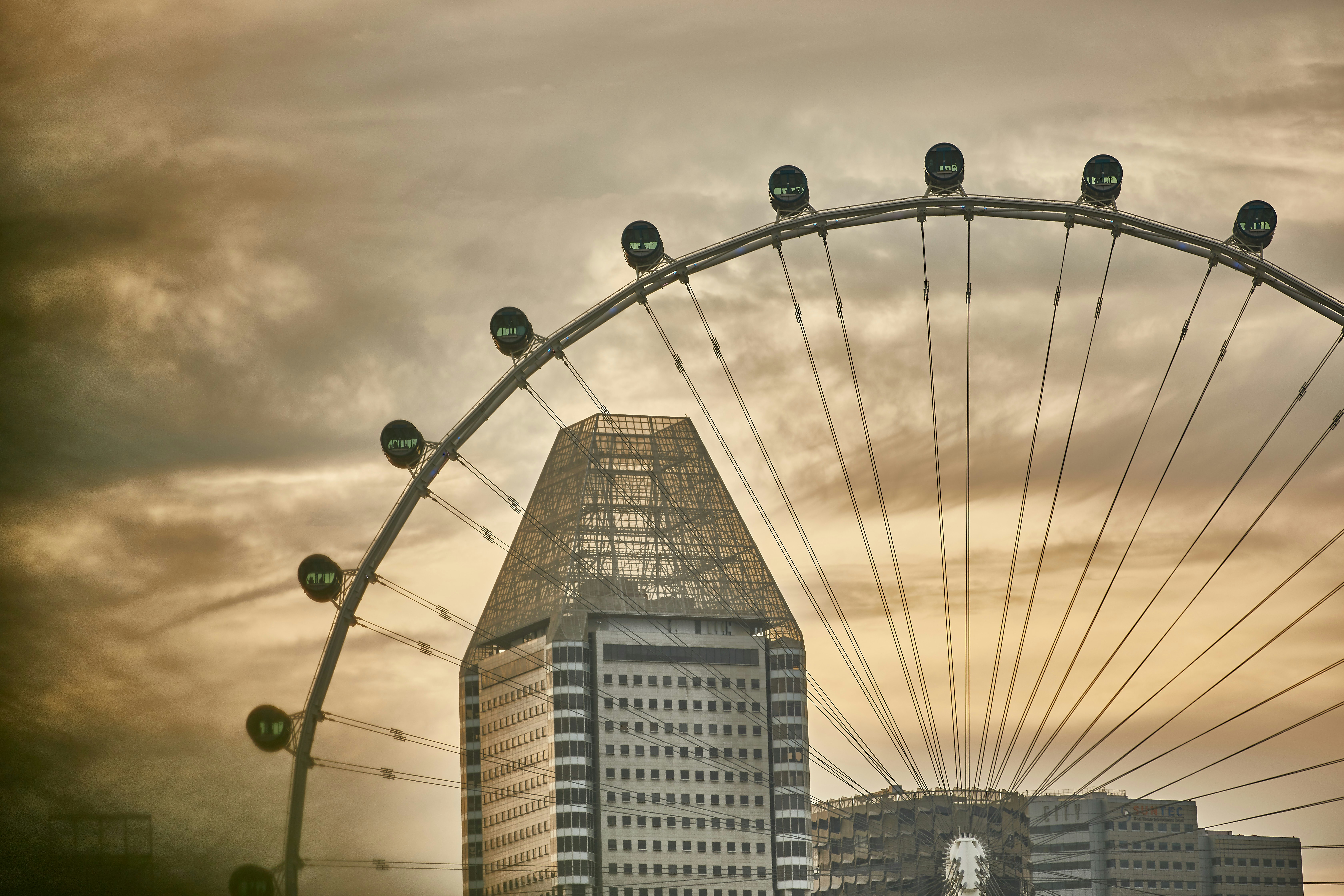 A modern Ferris wheel looms over a contemporary skyscraper against a dramatic sky. The architectural juxtaposition highlights the fusion of nature and urban design.