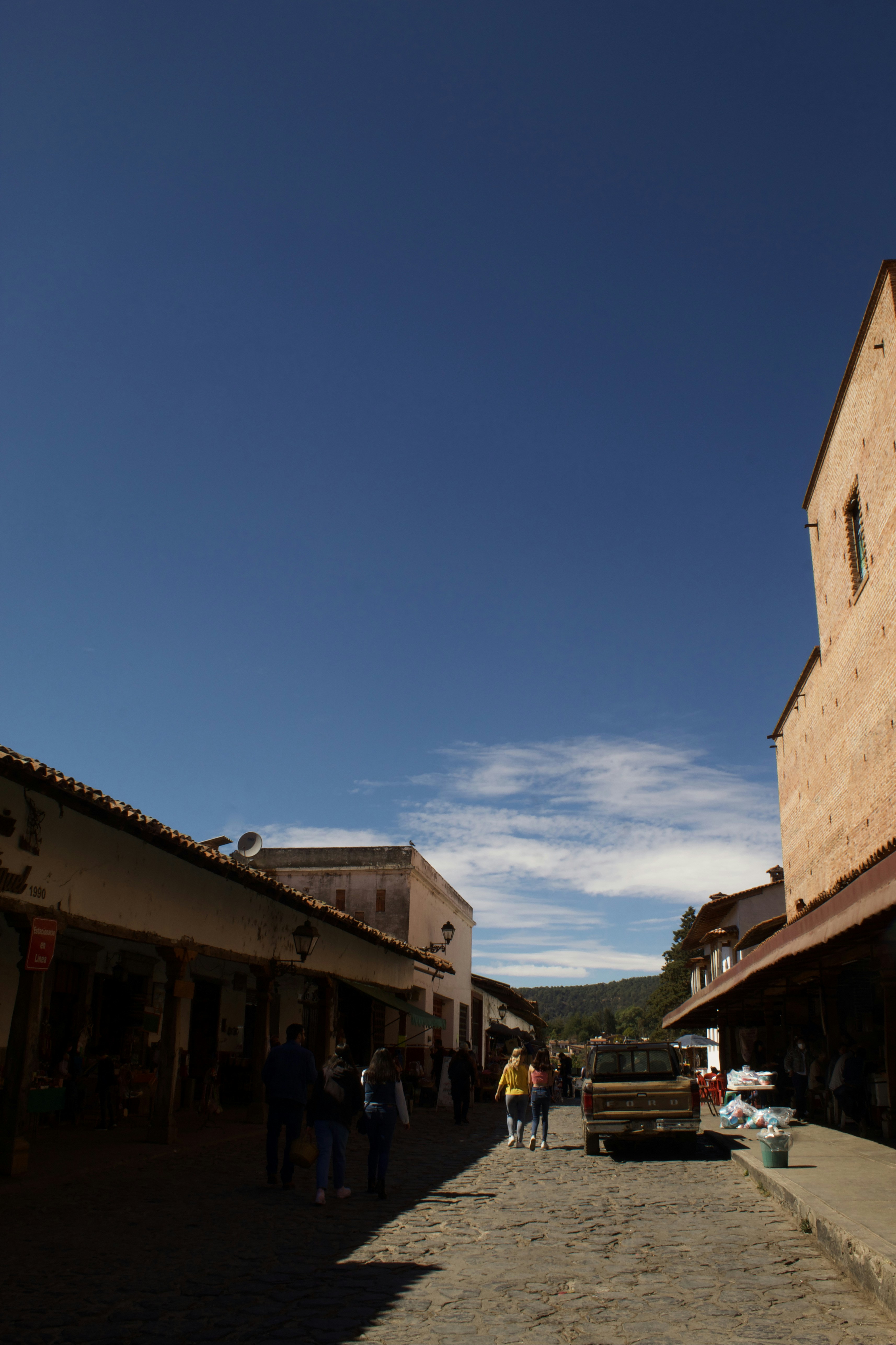 Historic cobblestone street lined with shops and people, under a clear blue sky. A vintage vehicle adds character to the scene.
