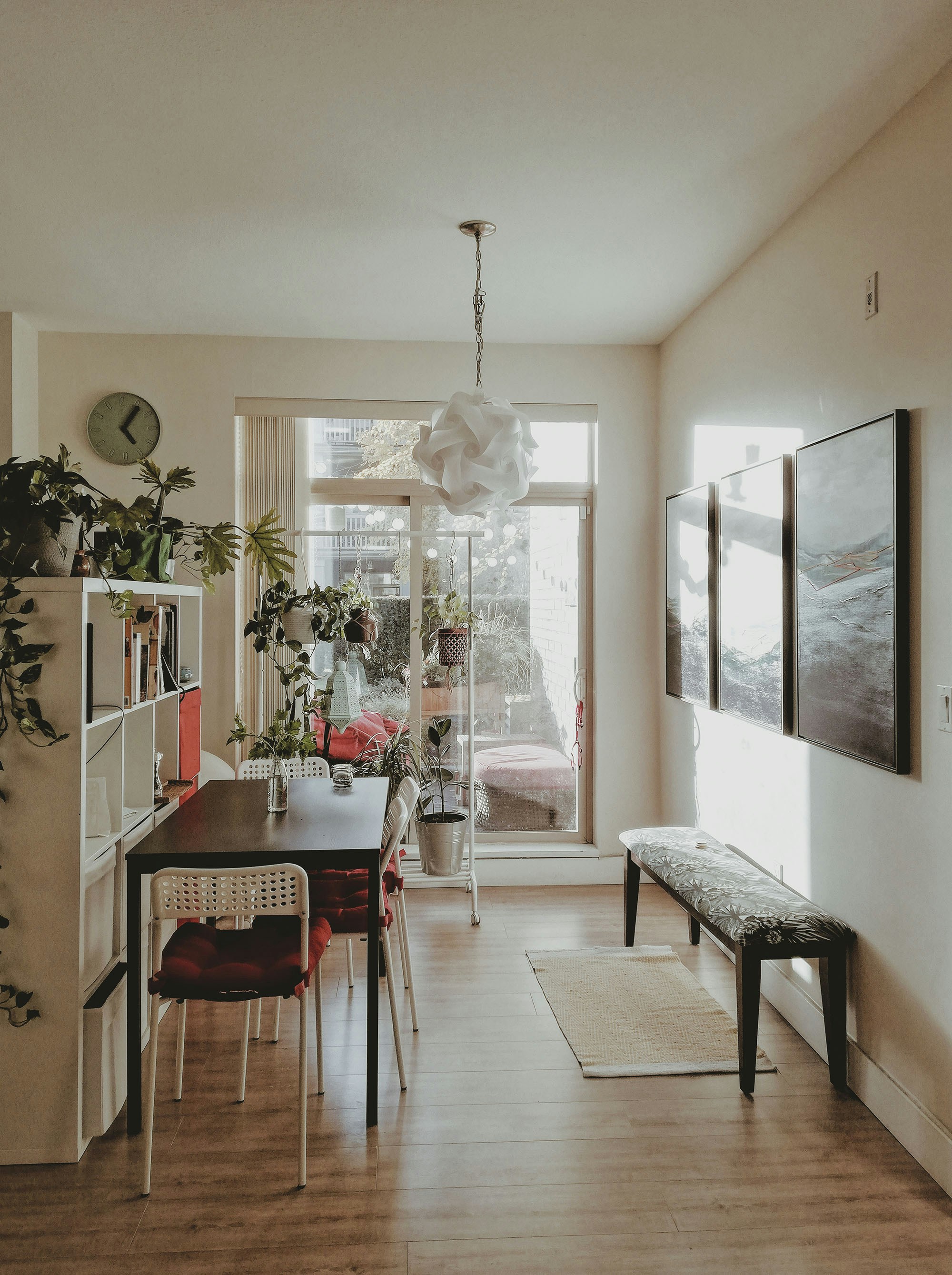 Brightly lit dining area featuring a wooden table, colorful chairs, and potted plants, with large windows allowing natural light to flood the space.