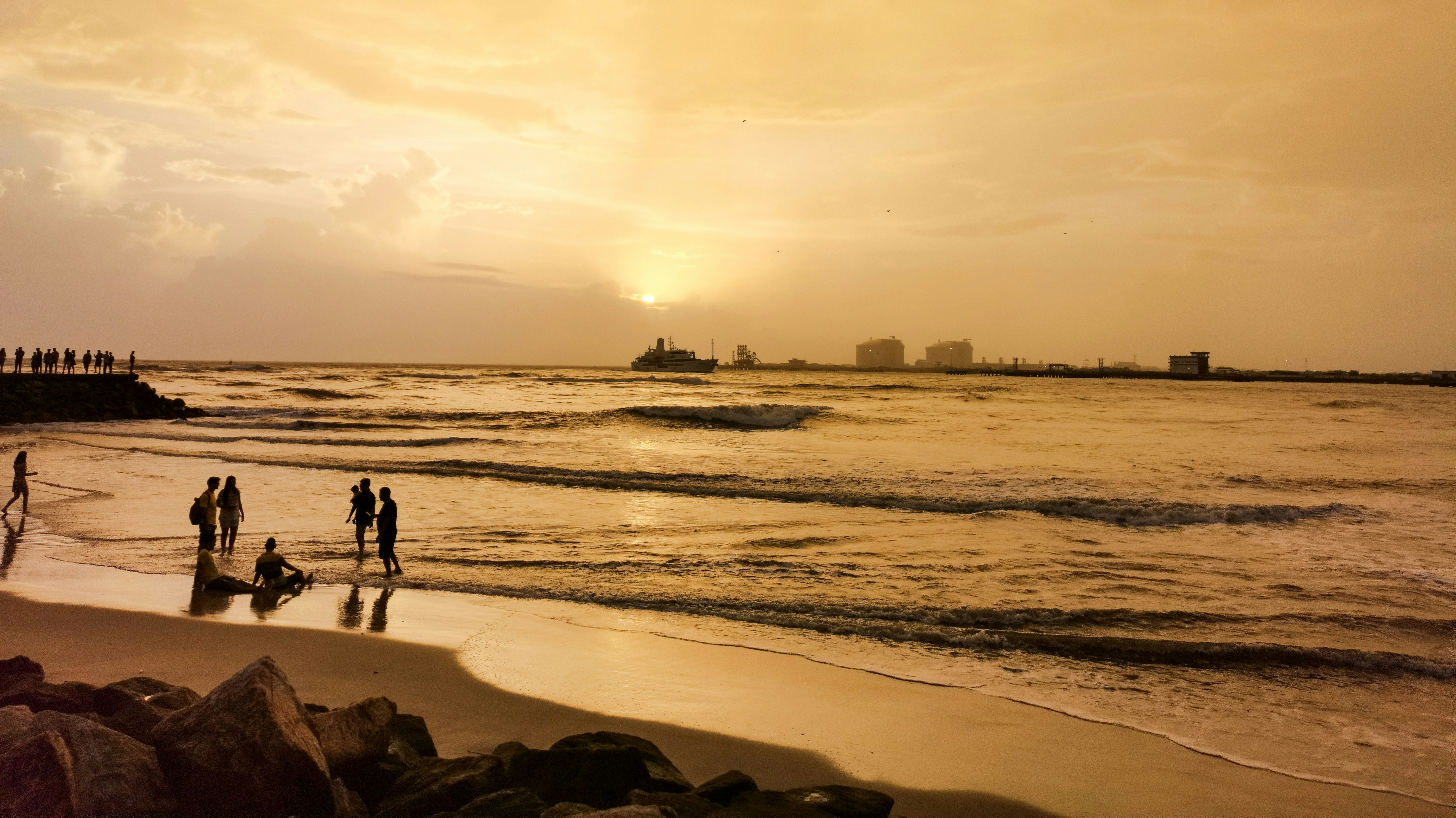 Golden sunset spills over a calm beach as silhouettes of beachgoers gather near the shore; distant buildings line the hazy horizon.