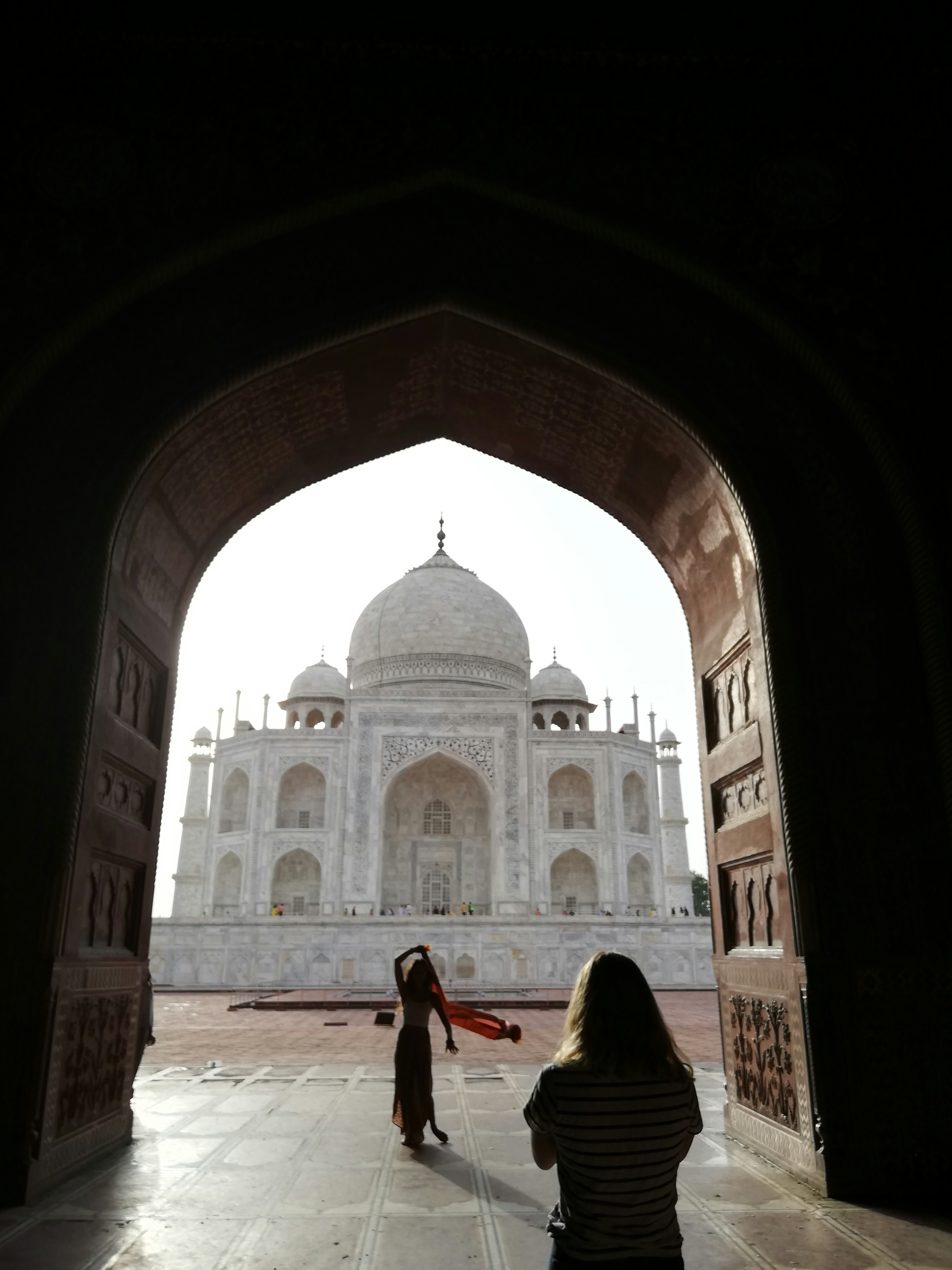 Photograph capturing the Taj Mahal framed by a dark arch. Silhouettes of two visitors stand in the foreground as the white mausoleum glows against the bright exterior.
