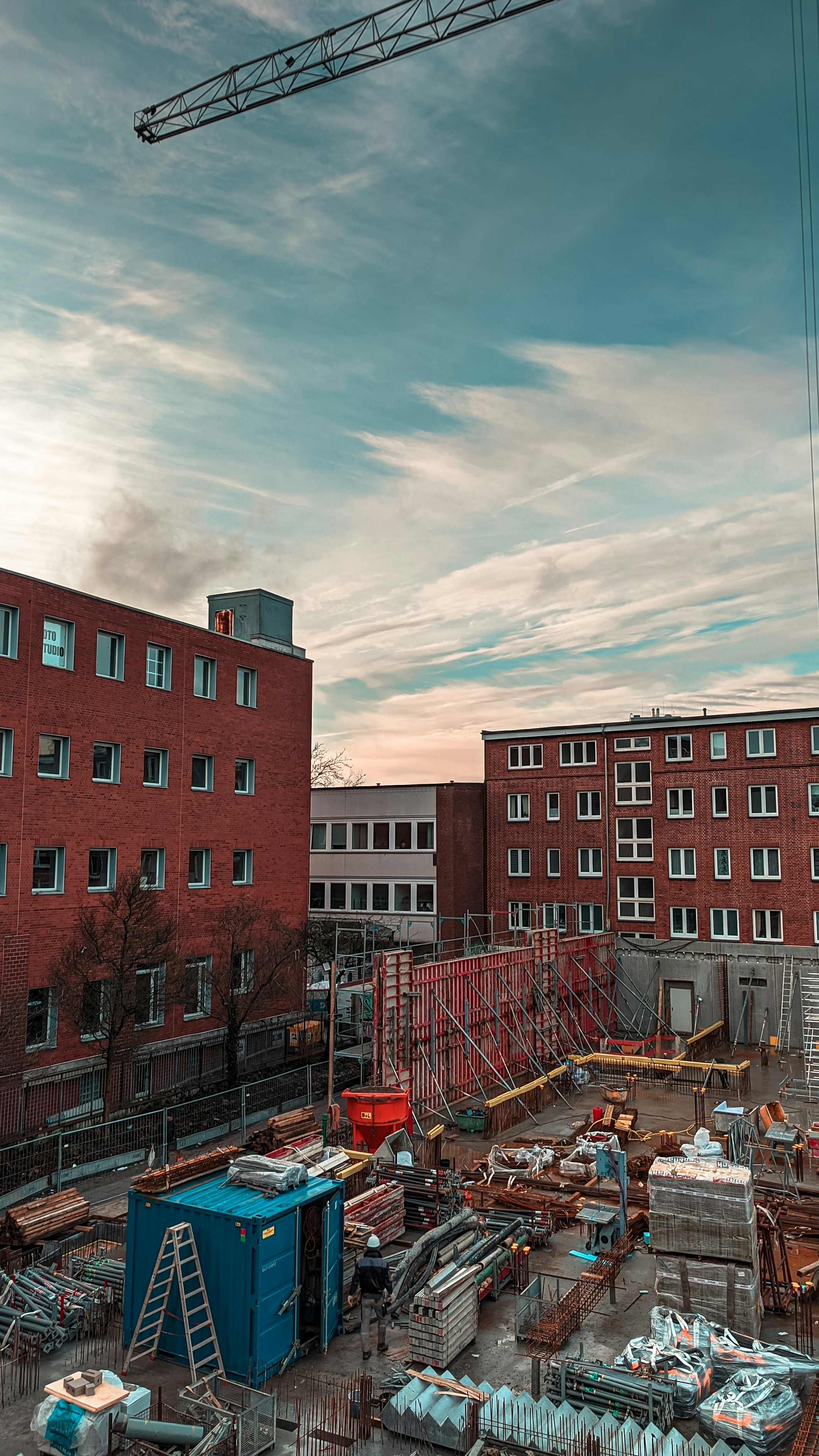 Construction site with red brick buildings and stacked materials under a blue, cloudy sky; a crane overhangs the frame as activity unfolds in the foreground.