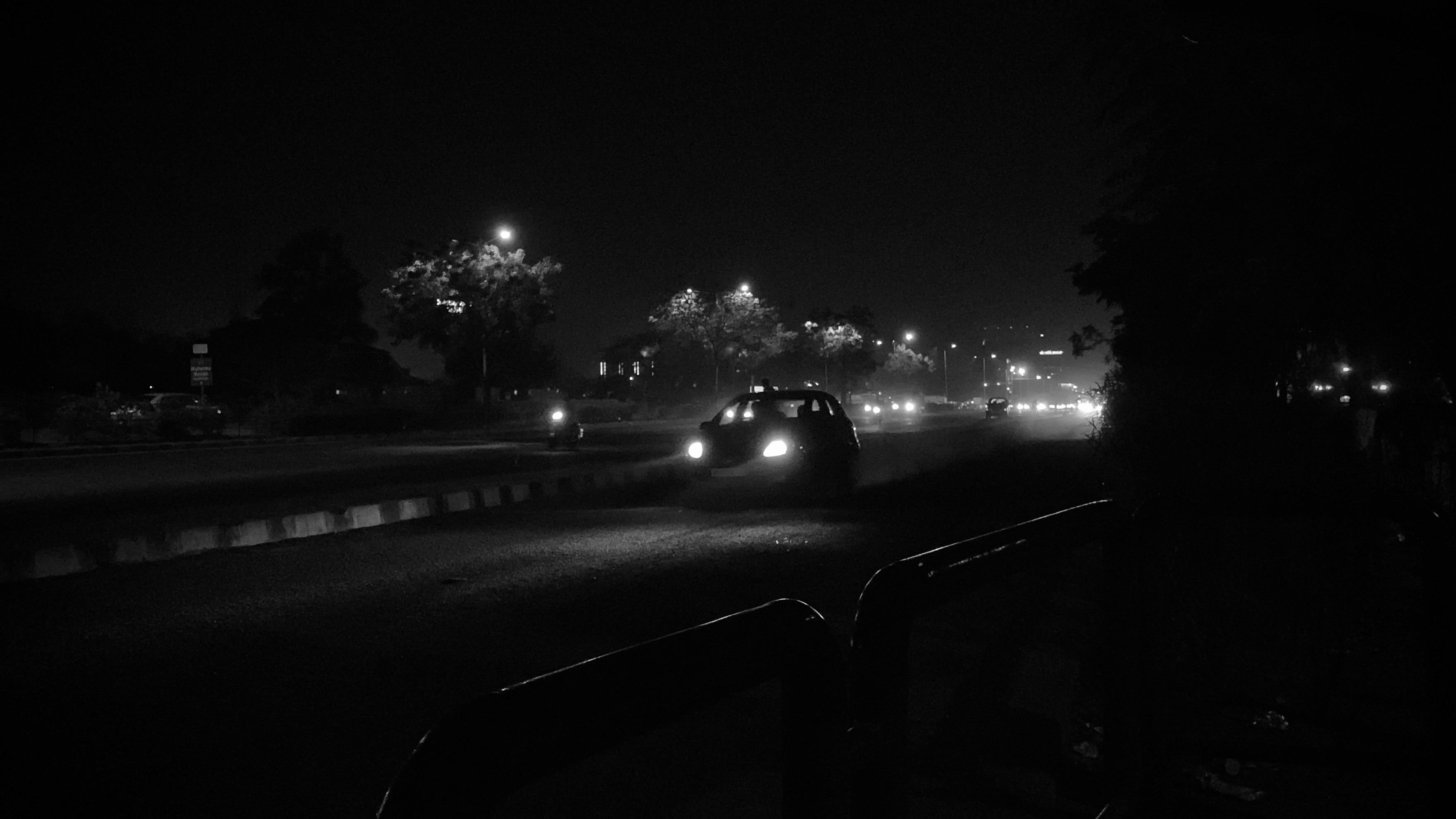 A solitary car navigates a dimly lit road, surrounded by faint streetlights and shadows of trees, creating a serene yet mysterious nighttime atmosphere.
