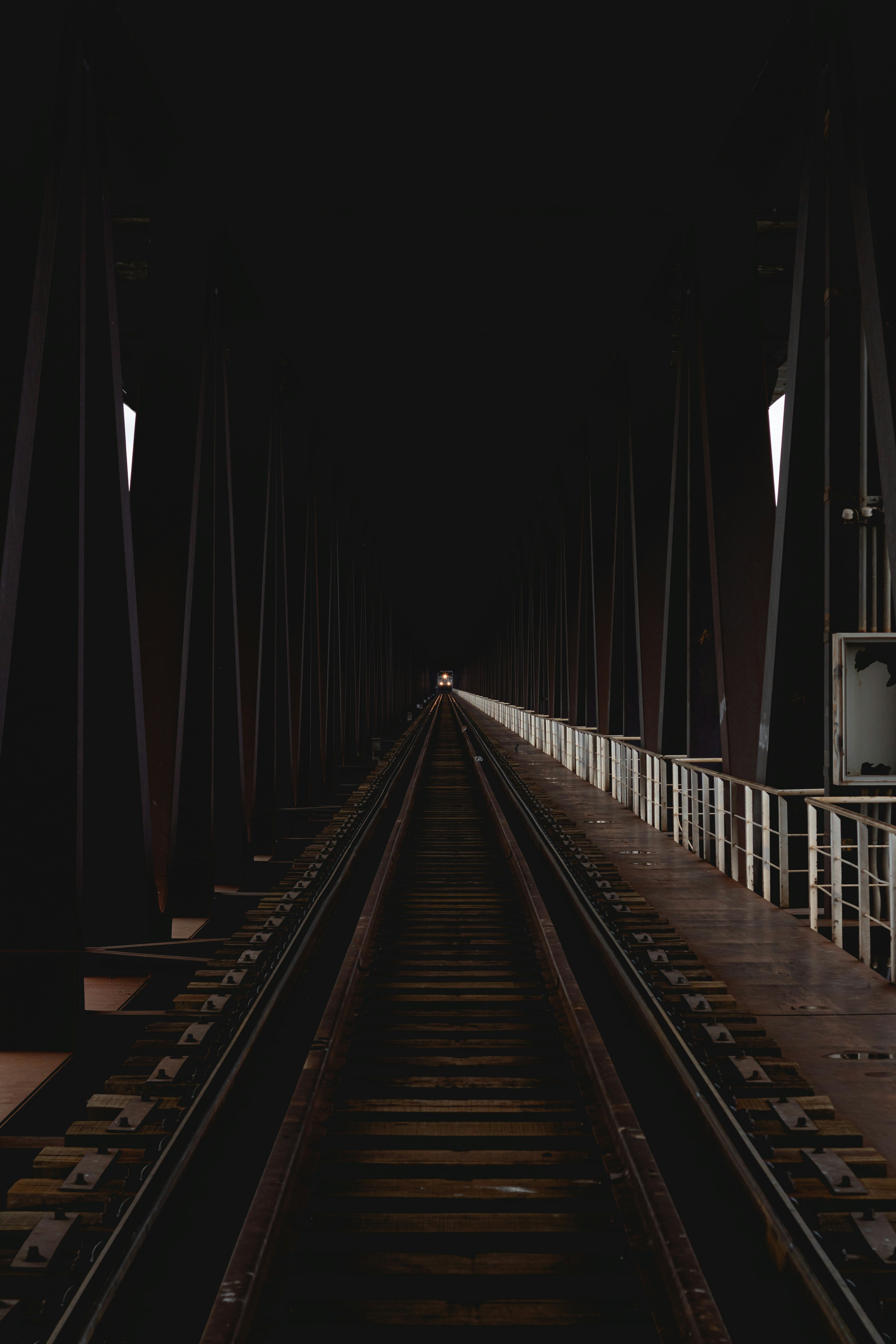 Railway tracks extending into a dark tunnel, framed by steel supports. The faint light at the end hints at an unseen destination.