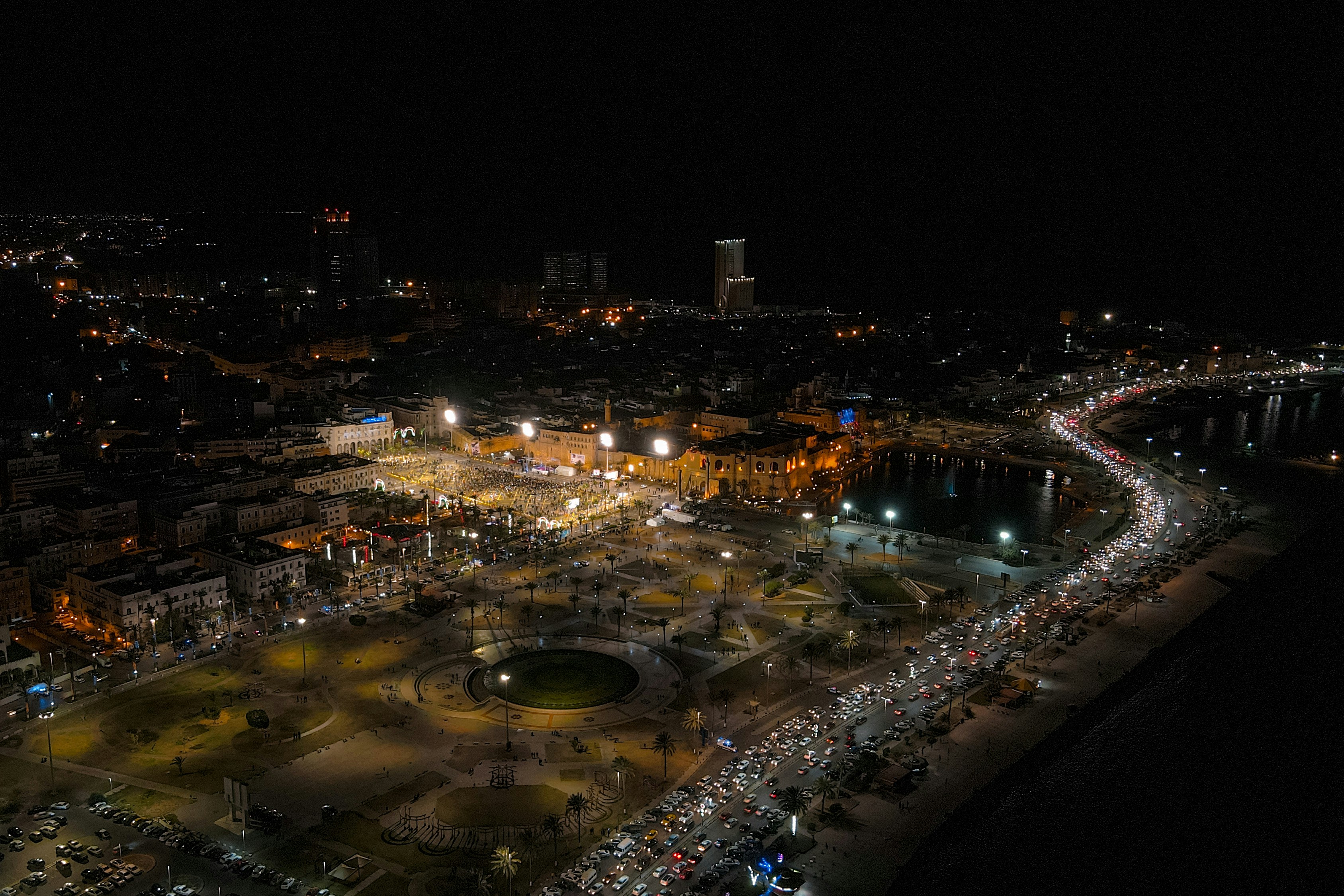 Aerial view of Tripoli's city lights and crowd gatherings at night along the coastline.