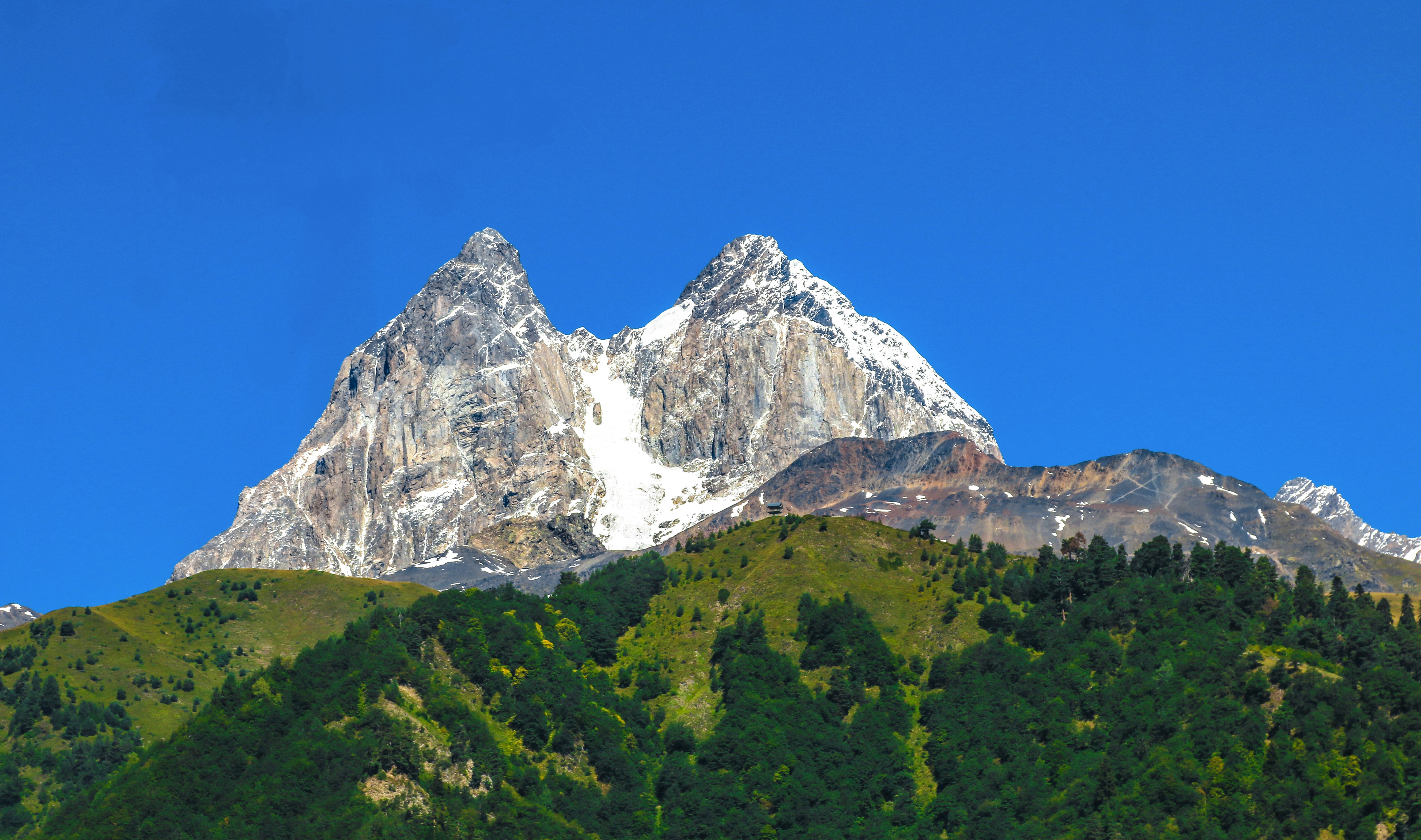 Snow-dusted twin mountain peaks rise above lush green forests under a vibrant blue sky.