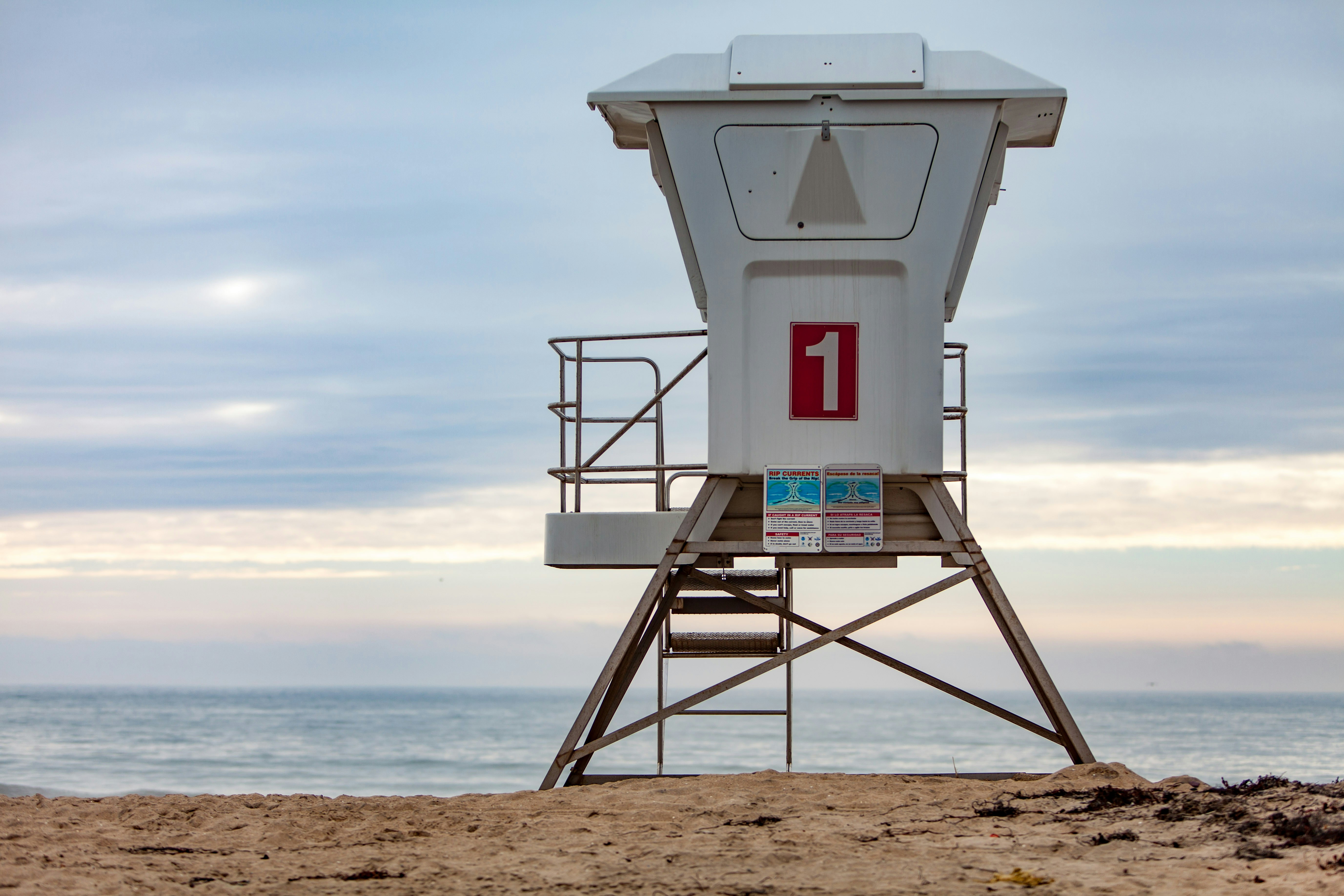 Lifeguard tower stands alone on a sandy beach with a calm ocean backdrop under a cloudy sky.