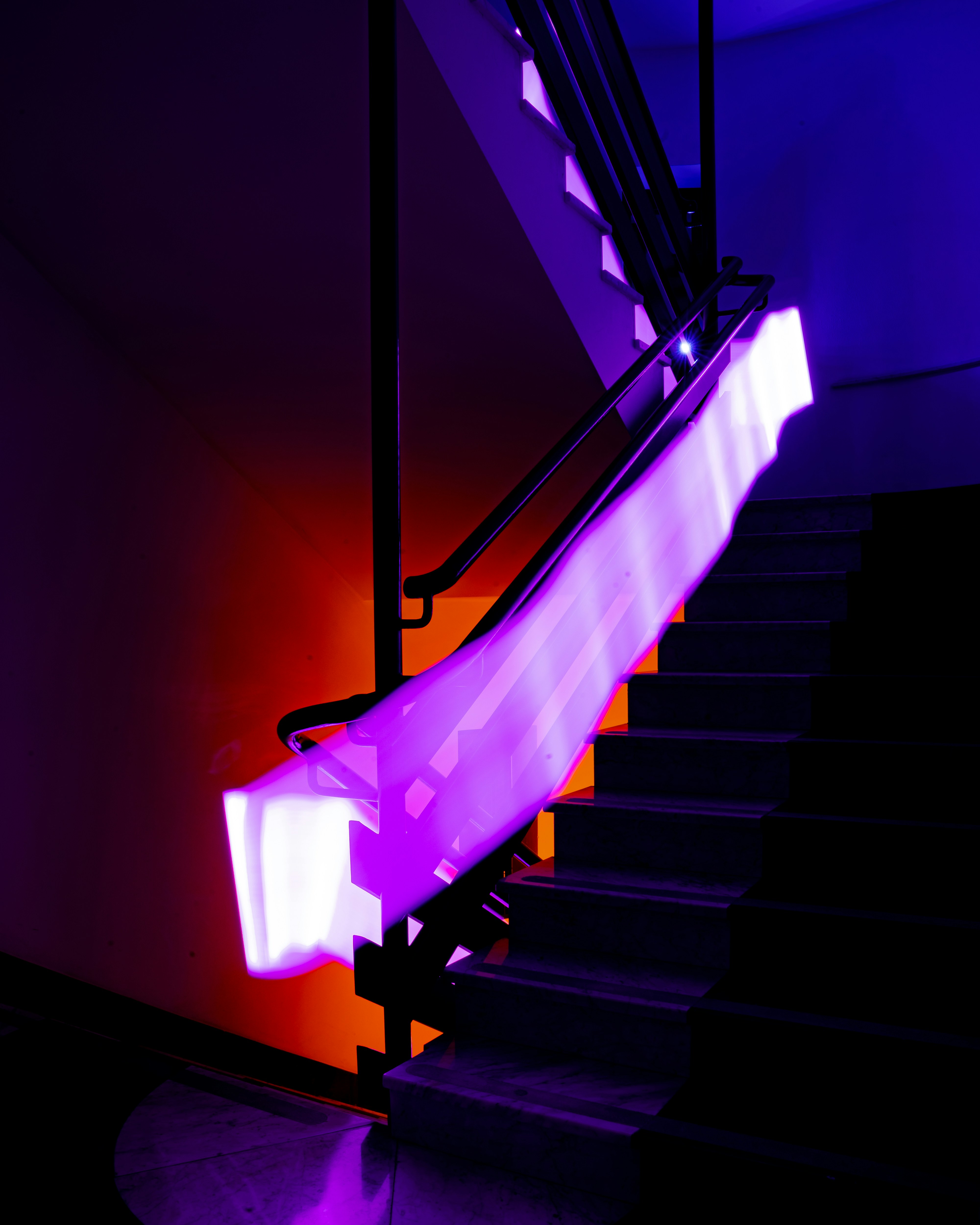 Vibrant purple light illuminating a staircase, creating a striking contrast with the surrounding dark walls. The interplay of colors guides the viewer's eye upward.