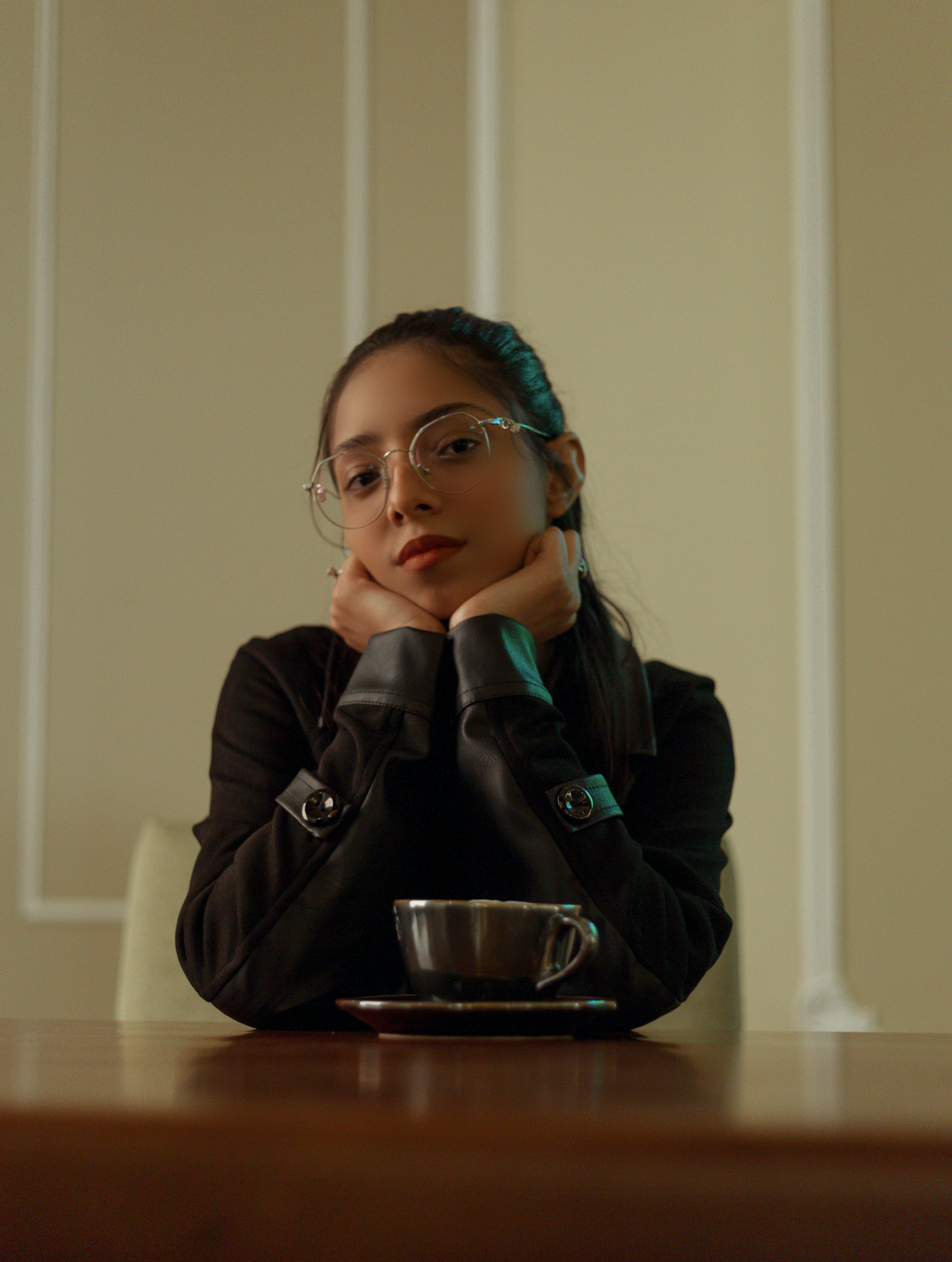 Young woman with glasses resting her chin on her hands, seated at a wooden table with a coffee cup and saucer in a warmly lit café.