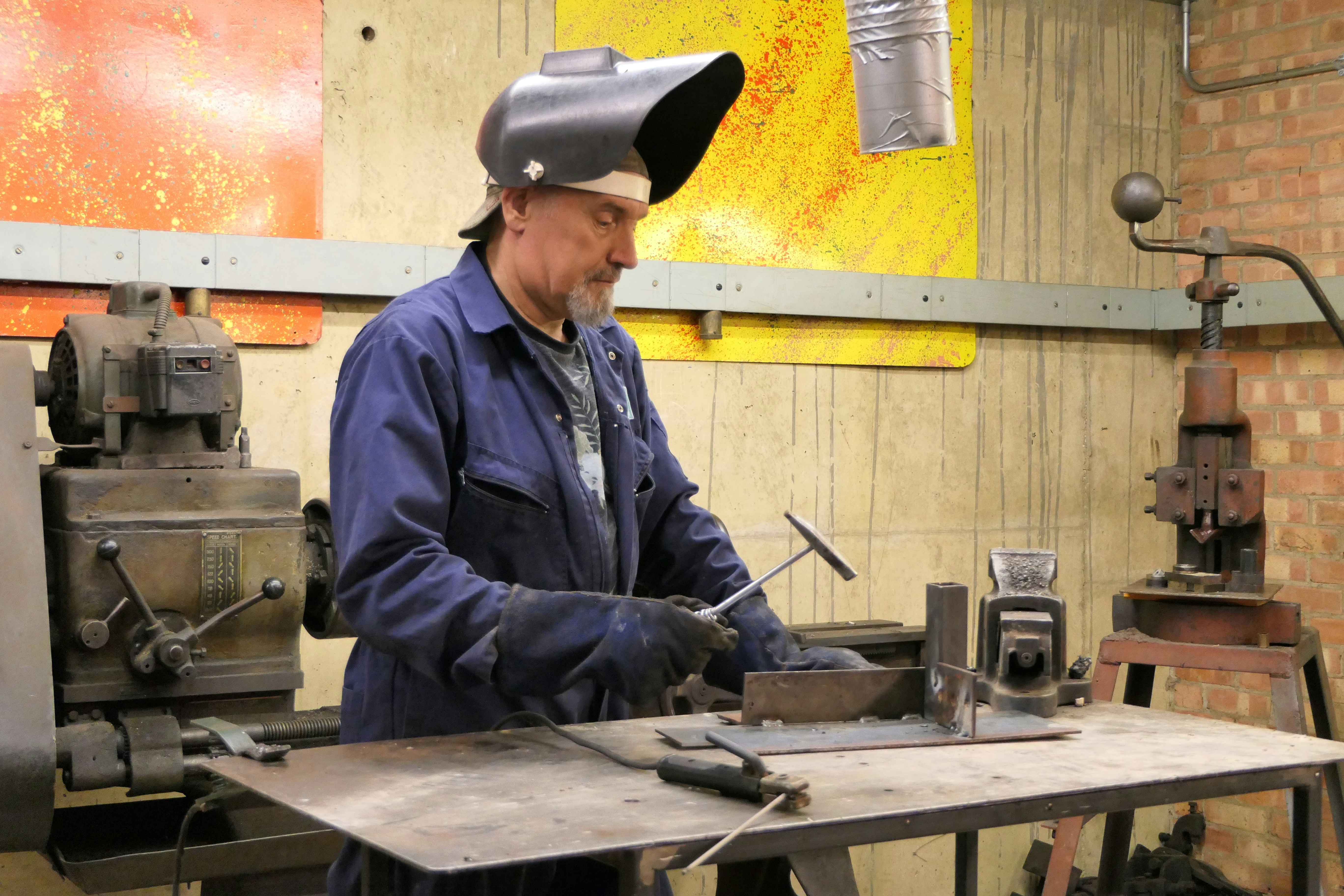Welder focused on fabricating metal components in a workshop filled with tools and vibrant artwork on the walls.