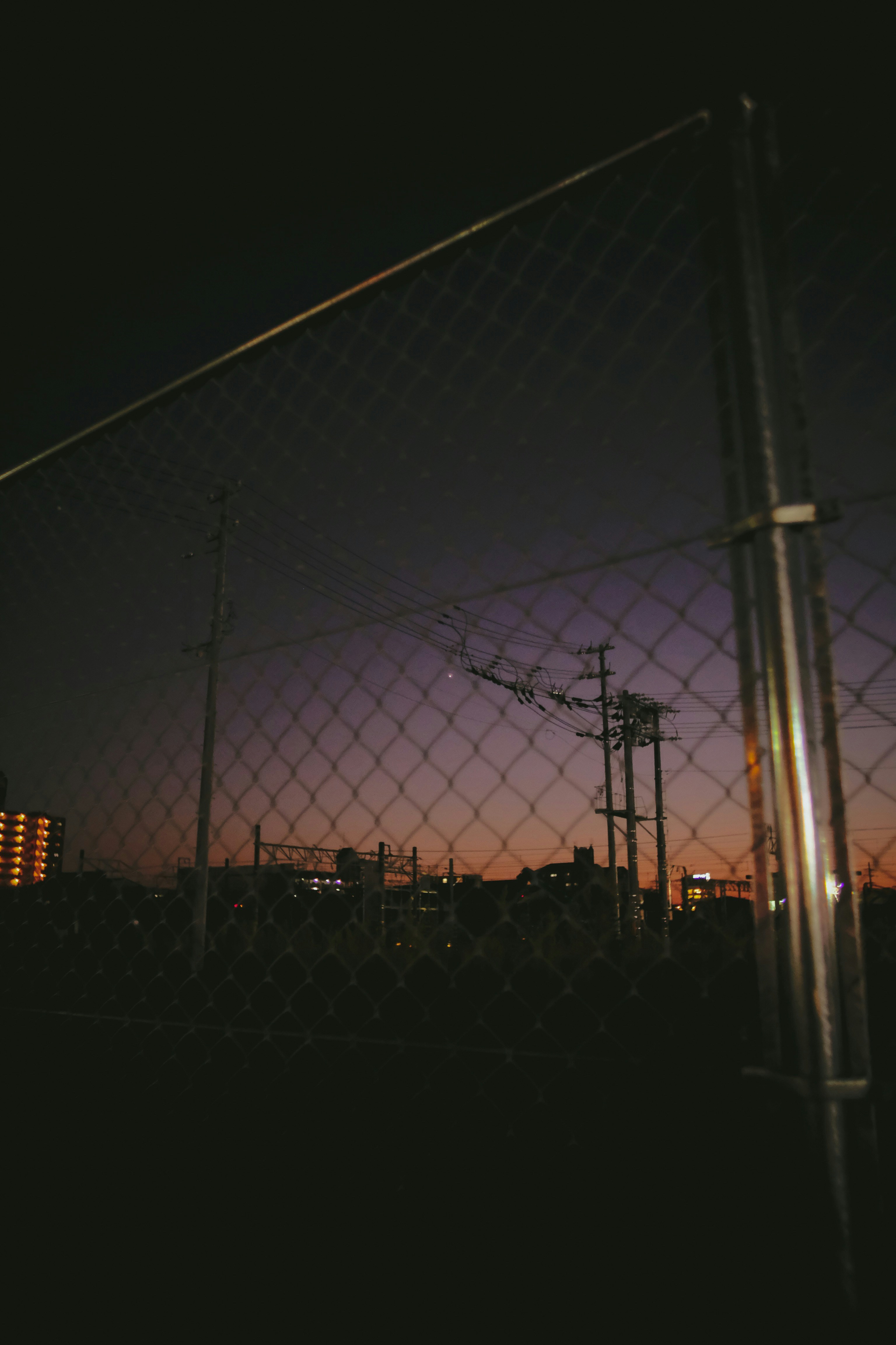 Silhouetted power lines and buildings framed by a chain-link fence at dusk, capturing the transition from day to night.