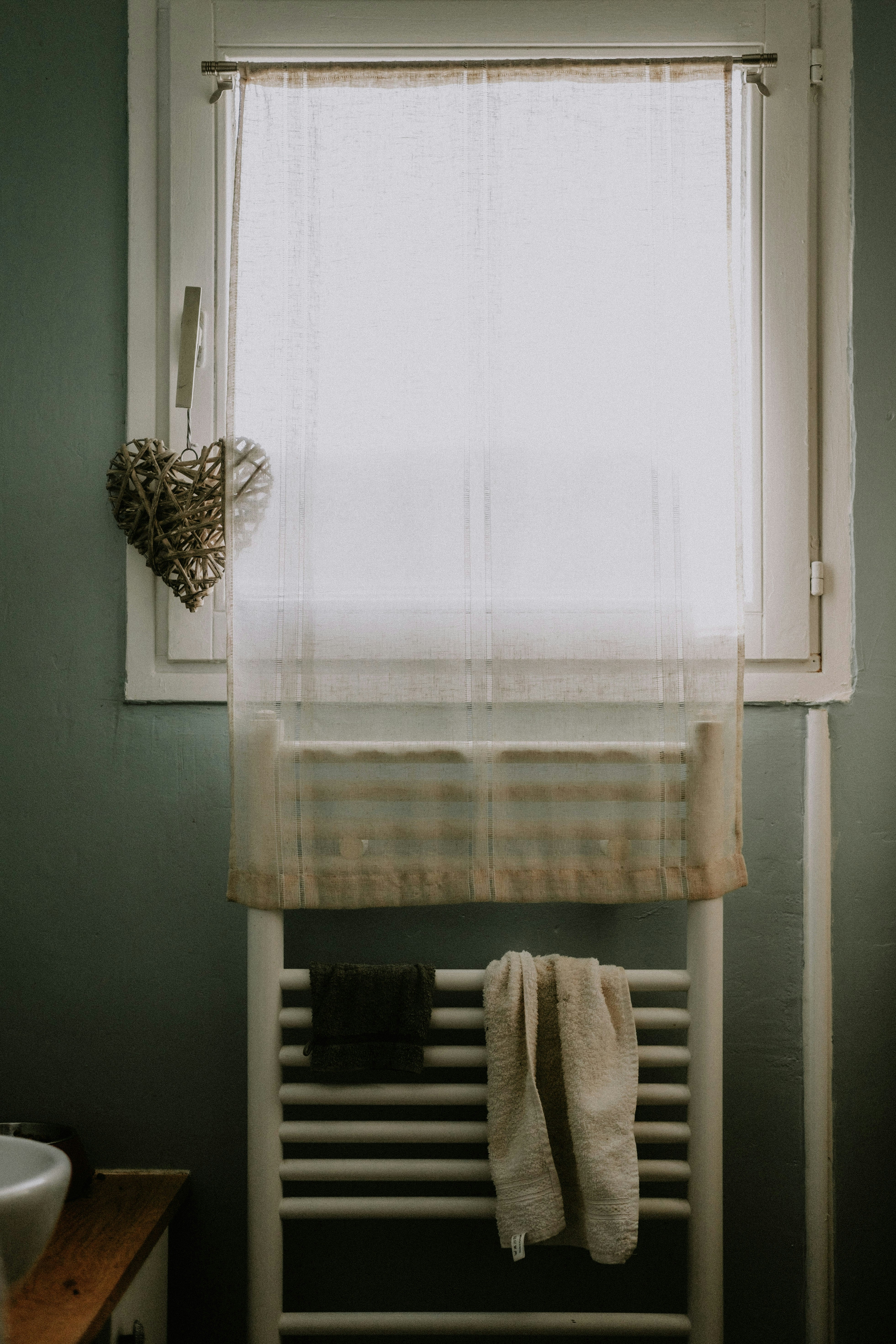 A heart-shaped decoration hangs near a softly lit window, complemented by neatly arranged towels on a white radiator. The calming green walls enhance the tranquil atmosphere.