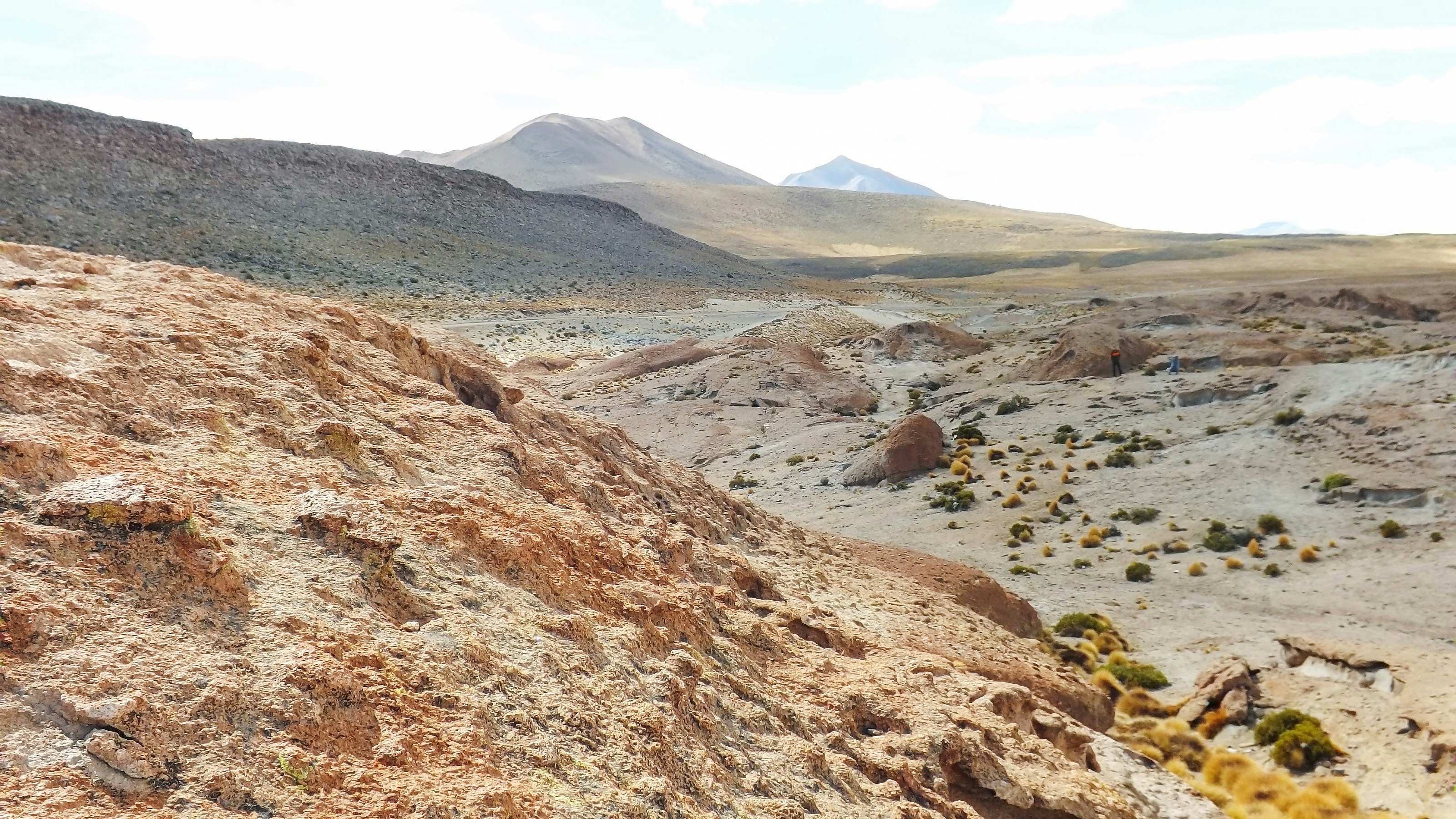 Vast arid terrain featuring rugged rock formations and distant mountains under a bright sky. The scene captures the essence of the high-altitude Bolivian landscape.