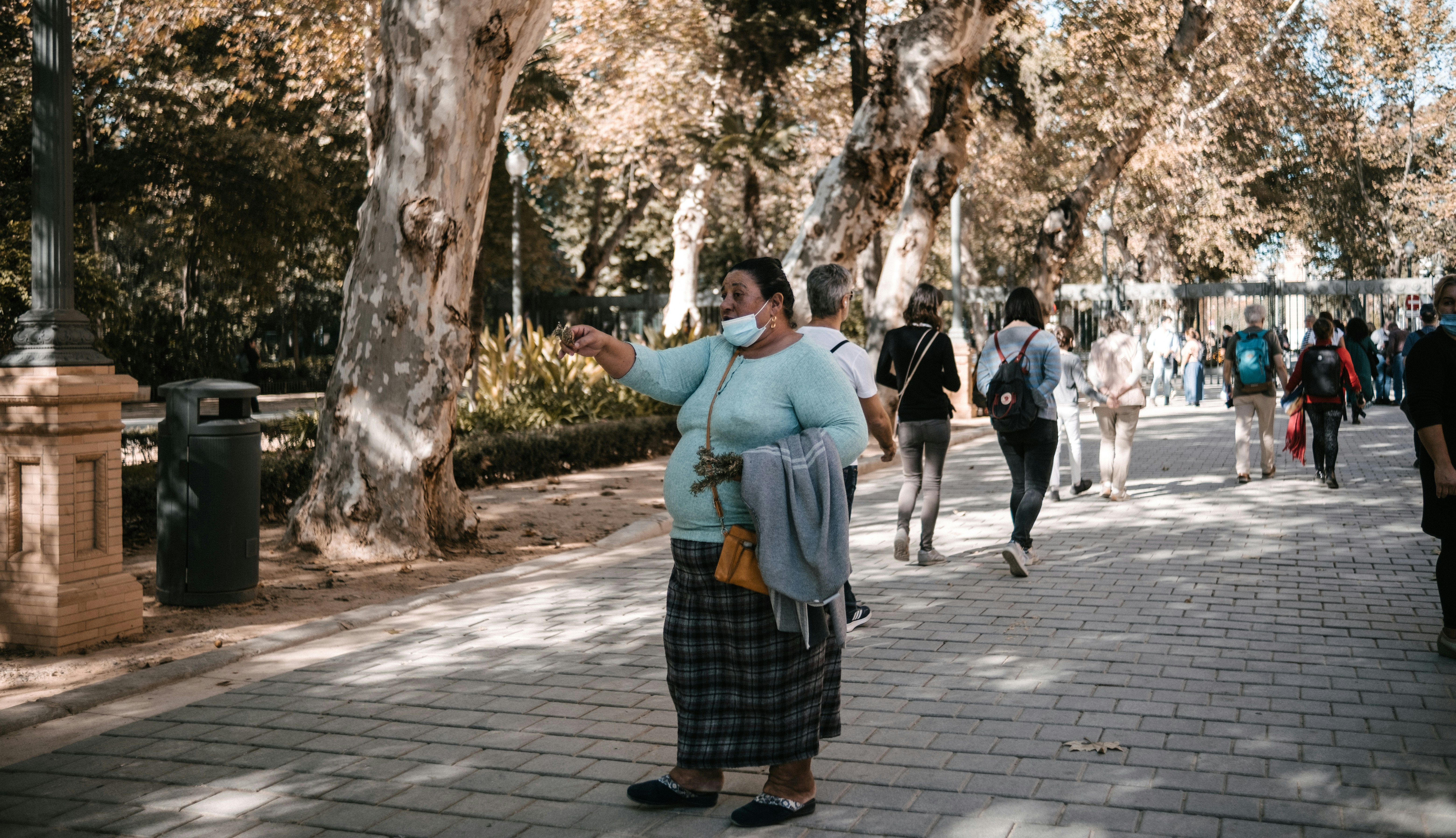 Woman in a mask gesturing on a tree-lined sidewalk amidst pedestrians.