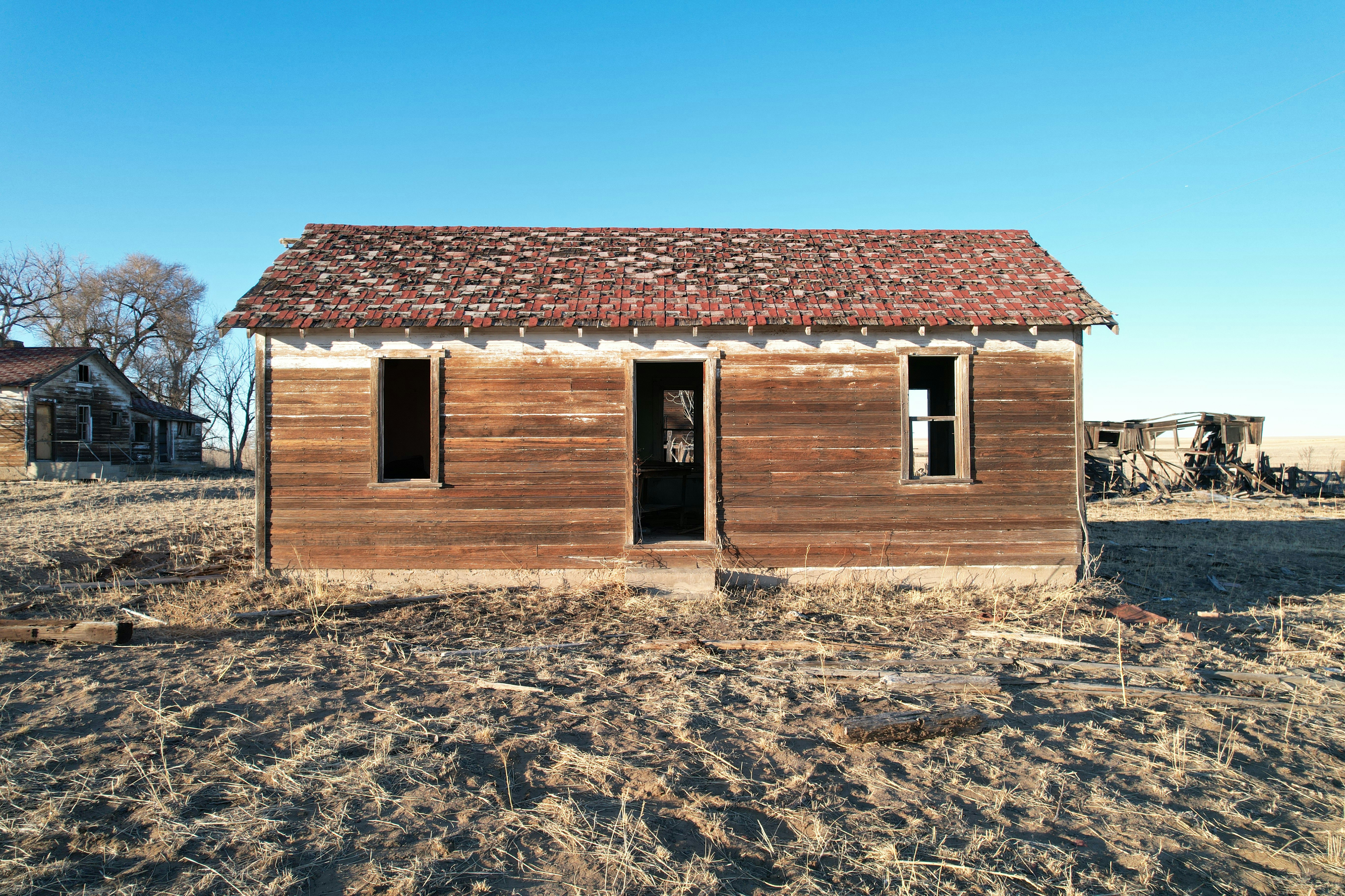 Abandoned wooden house with a weathered facade, standing in a dry landscape under a clear blue sky. The structure hints at a forgotten history.