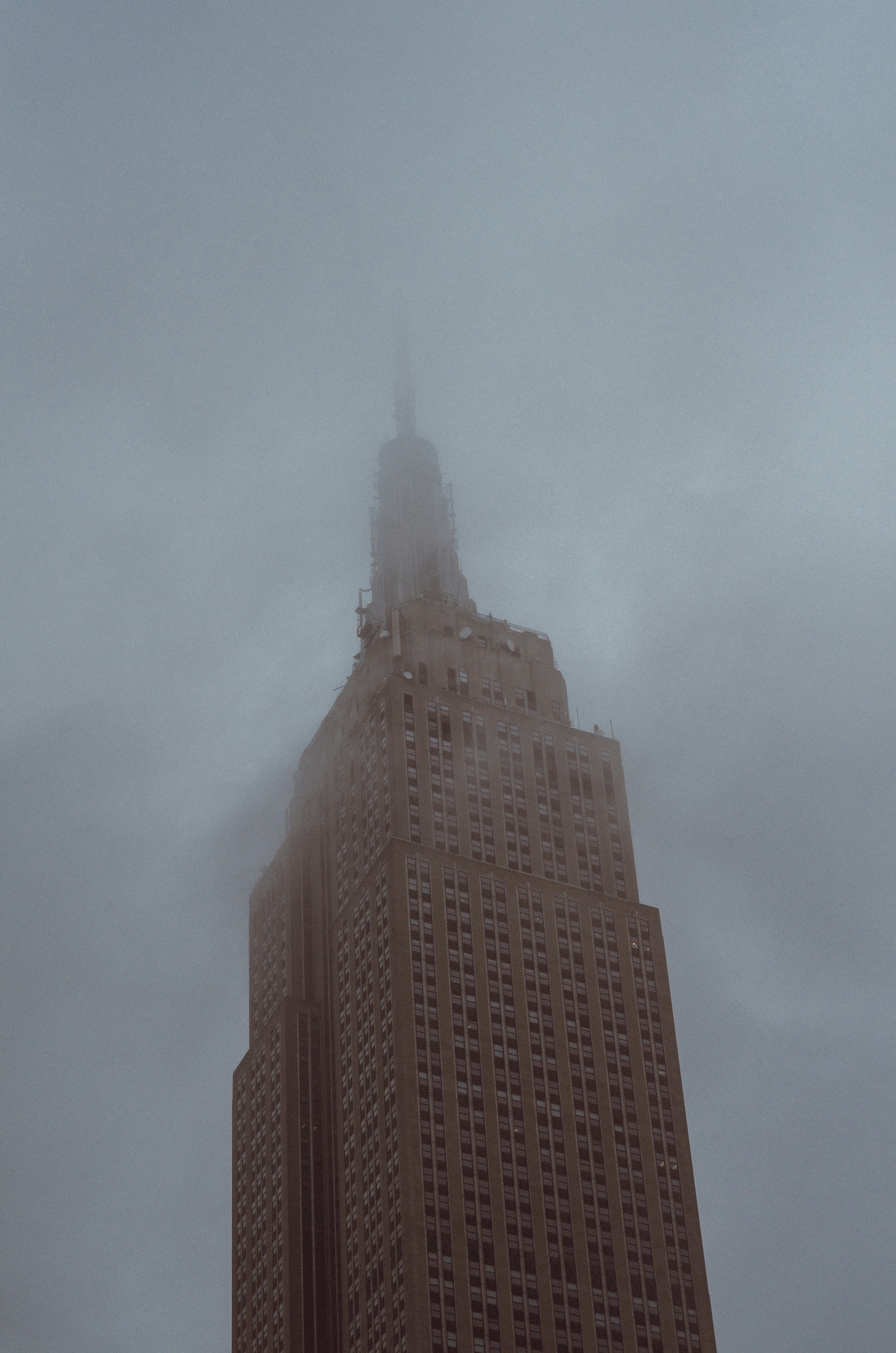 The Empire State Building rises through a dense fog, its iconic silhouette partially obscured by clouds. A striking representation of urban architecture in atmospheric conditions.