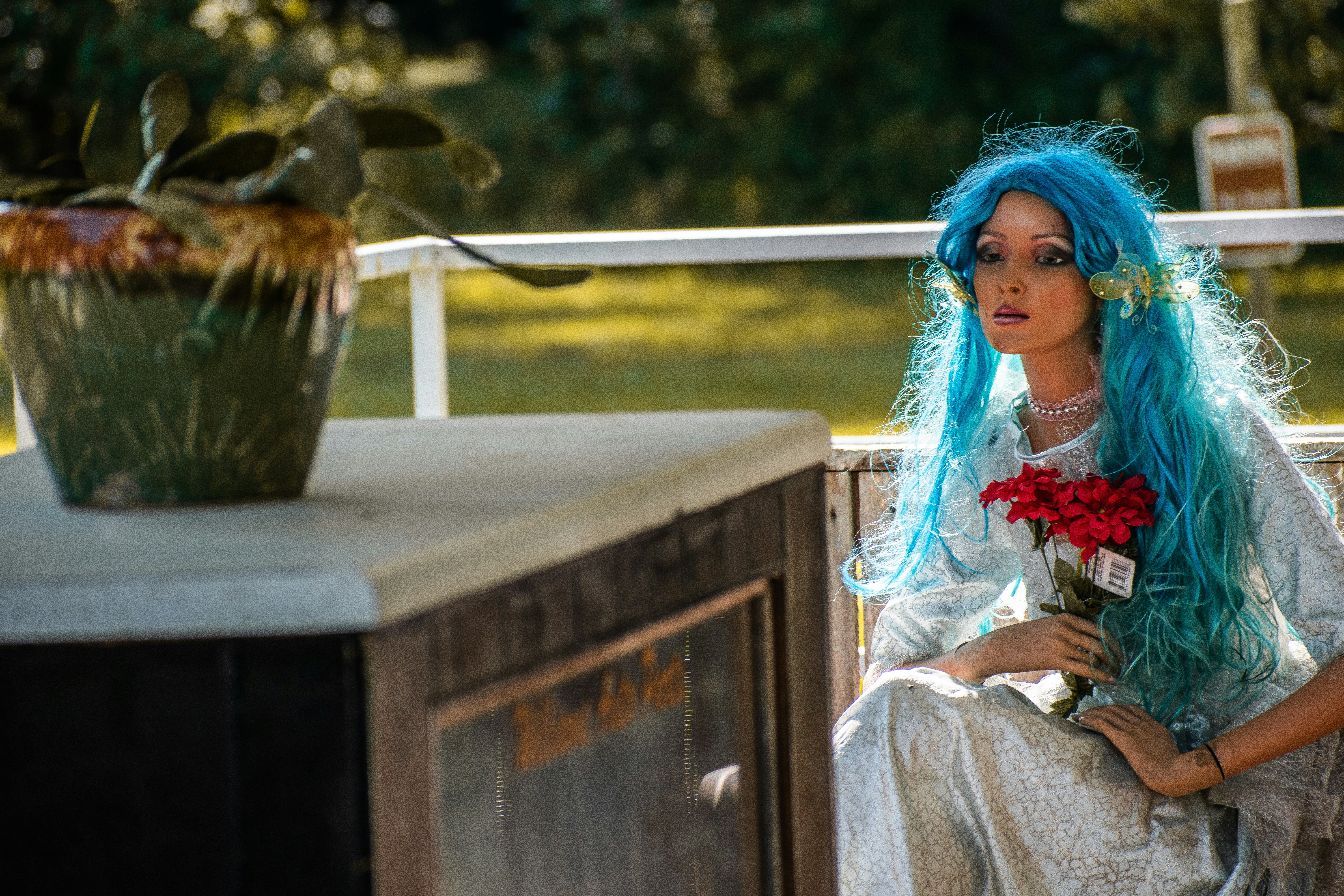 A serene figure with vibrant blue hair holds a bouquet of red flowers, seated beside a potted plant in a sunlit outdoor setting.