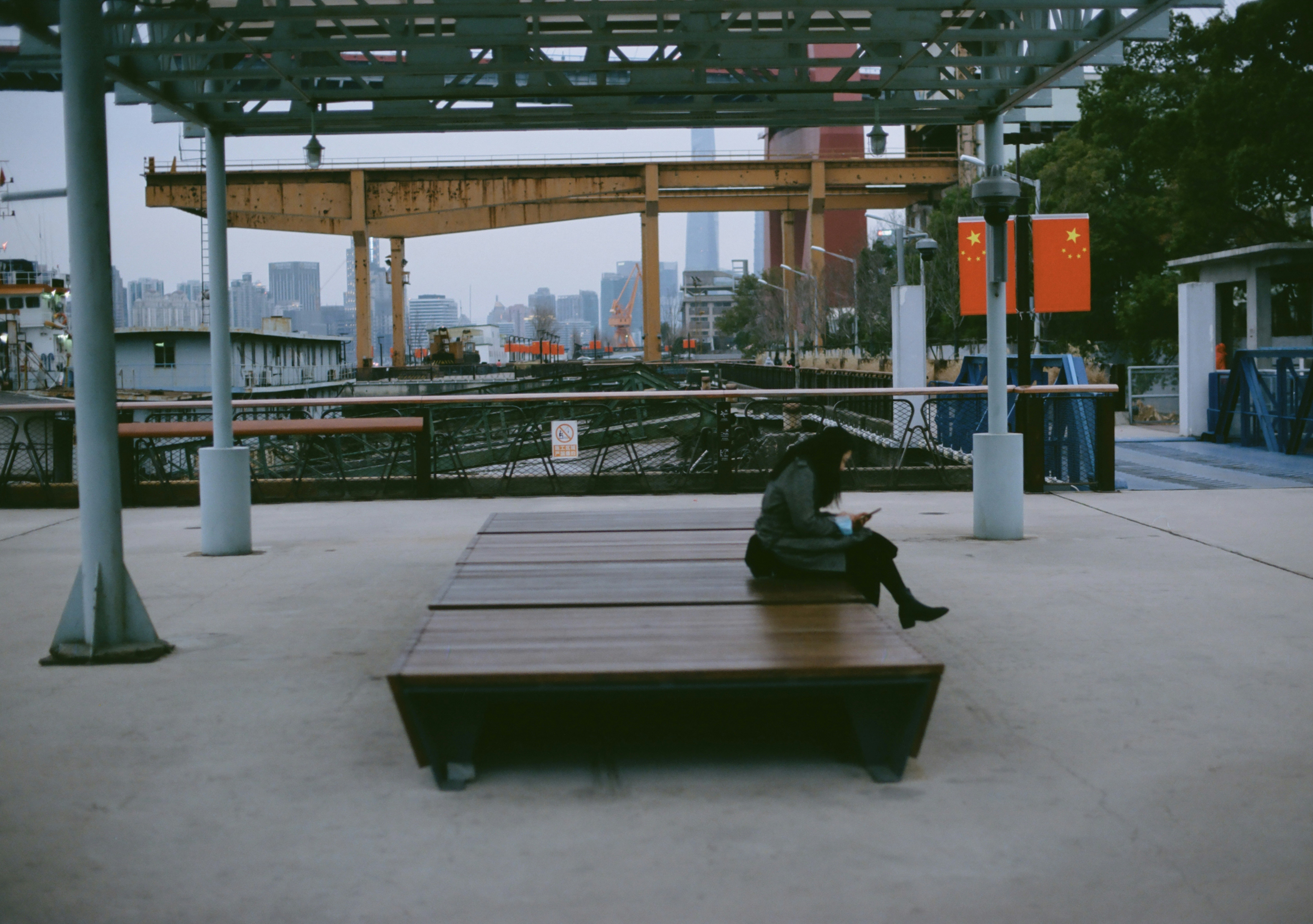 A solitary figure sits on a wooden bench in an urban setting, surrounded by construction elements and flags, reflecting a moment of quiet amidst bustling activity.